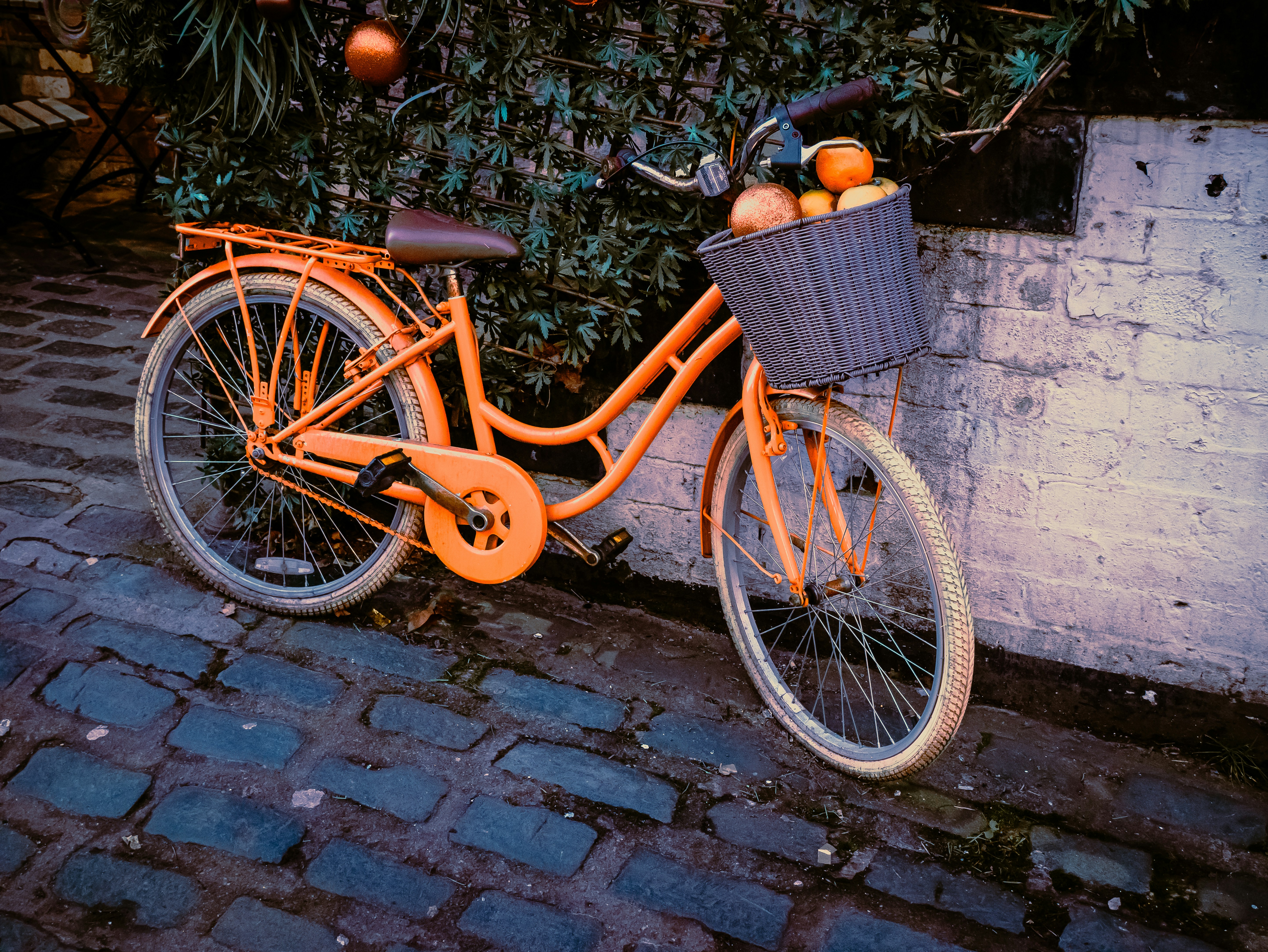 An orange-painted bicycle with a front basket of oranges rests against a stone wall on a cobblestone street, captured in a daytime photograph.