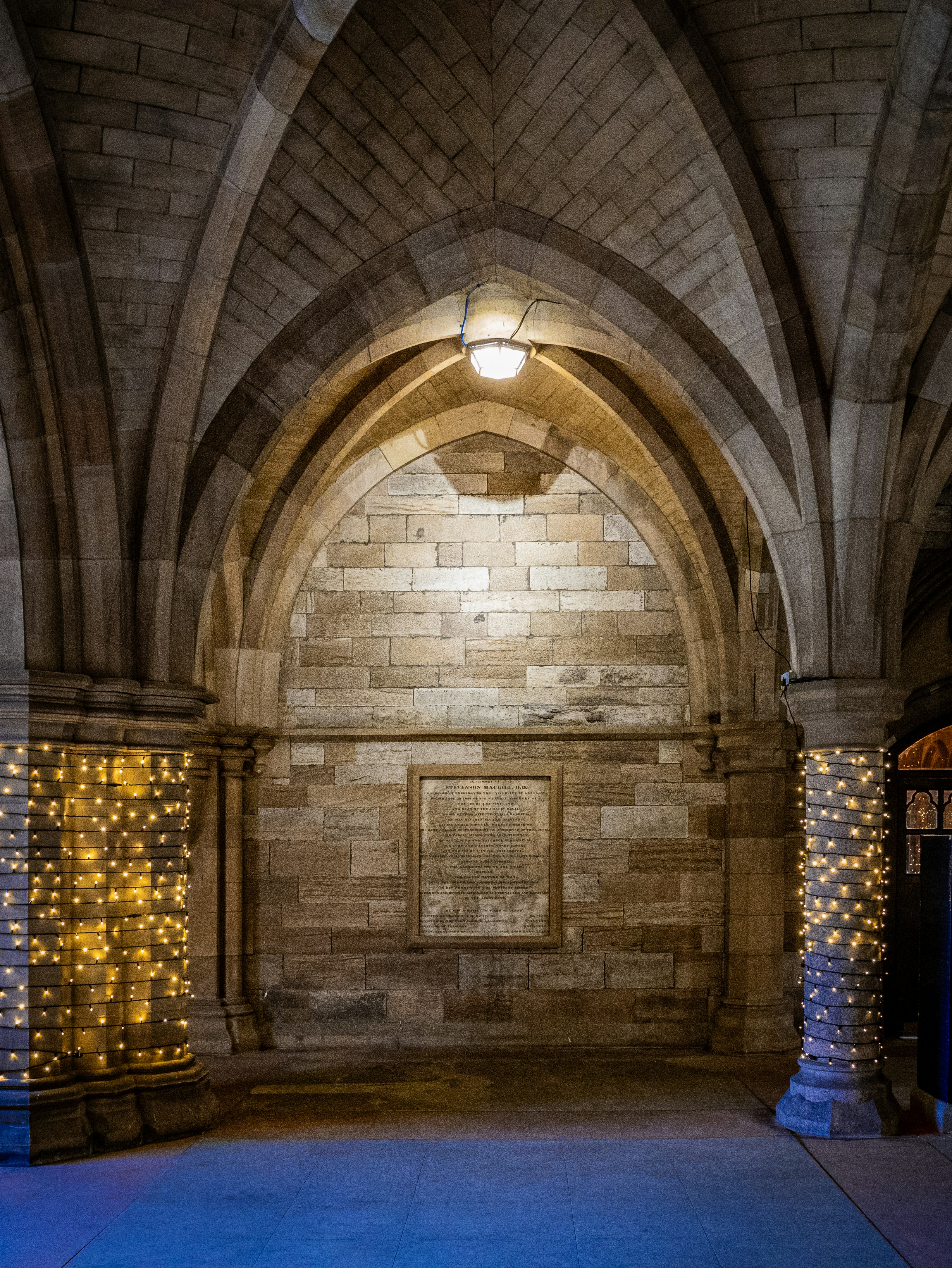 A serene alcove within the University of Glasgow's cloisters, where fairy lights gently embrace a column, creating a juxtaposition of warmth against the cool stone. A historical plaque mounted on the wall adds depth to the storied atmosphere of this tranquil corner of the campus.