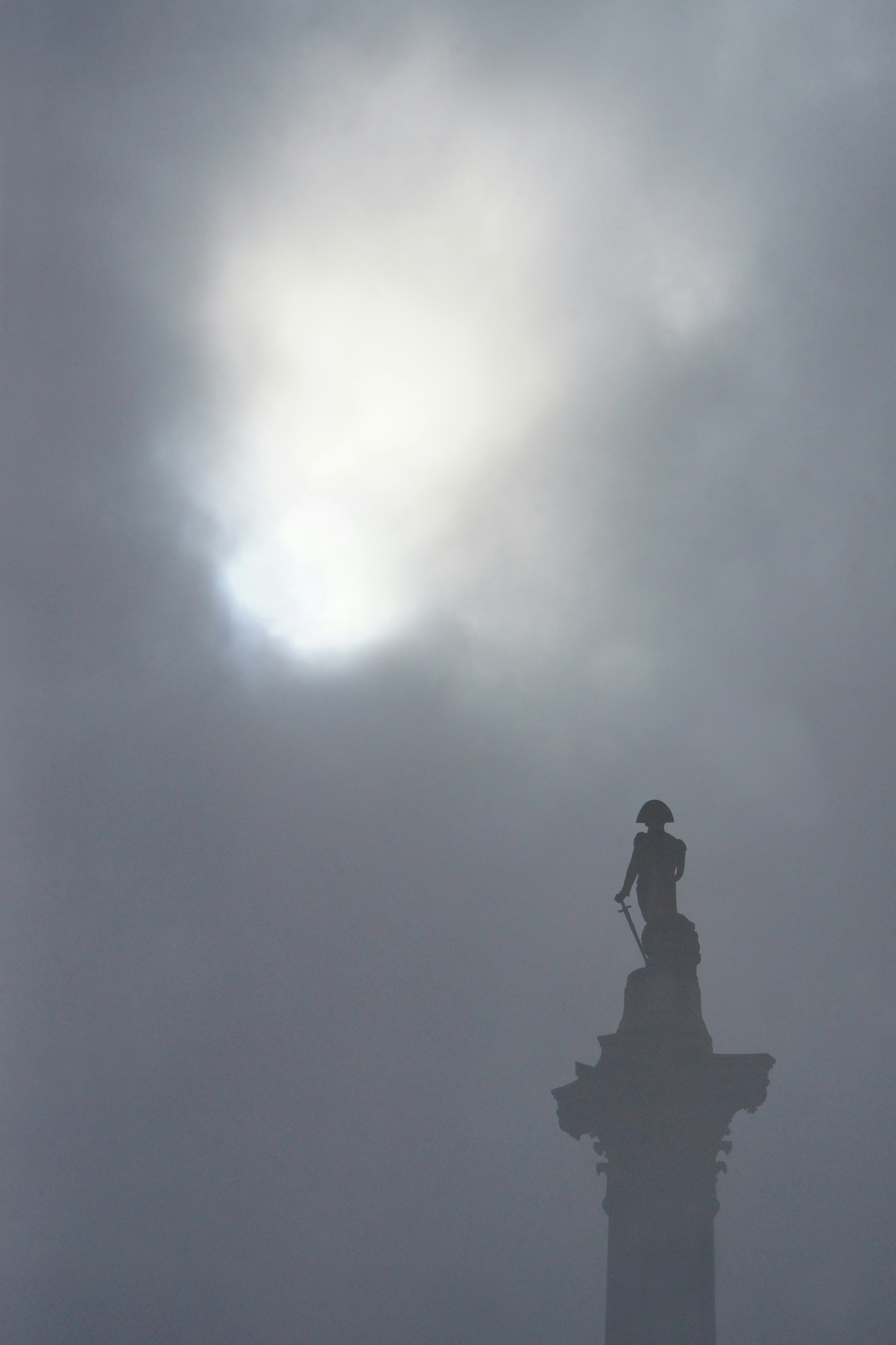 Silhouette of a statue atop a column, shrouded in fog with a faint light breaking through the clouds above.