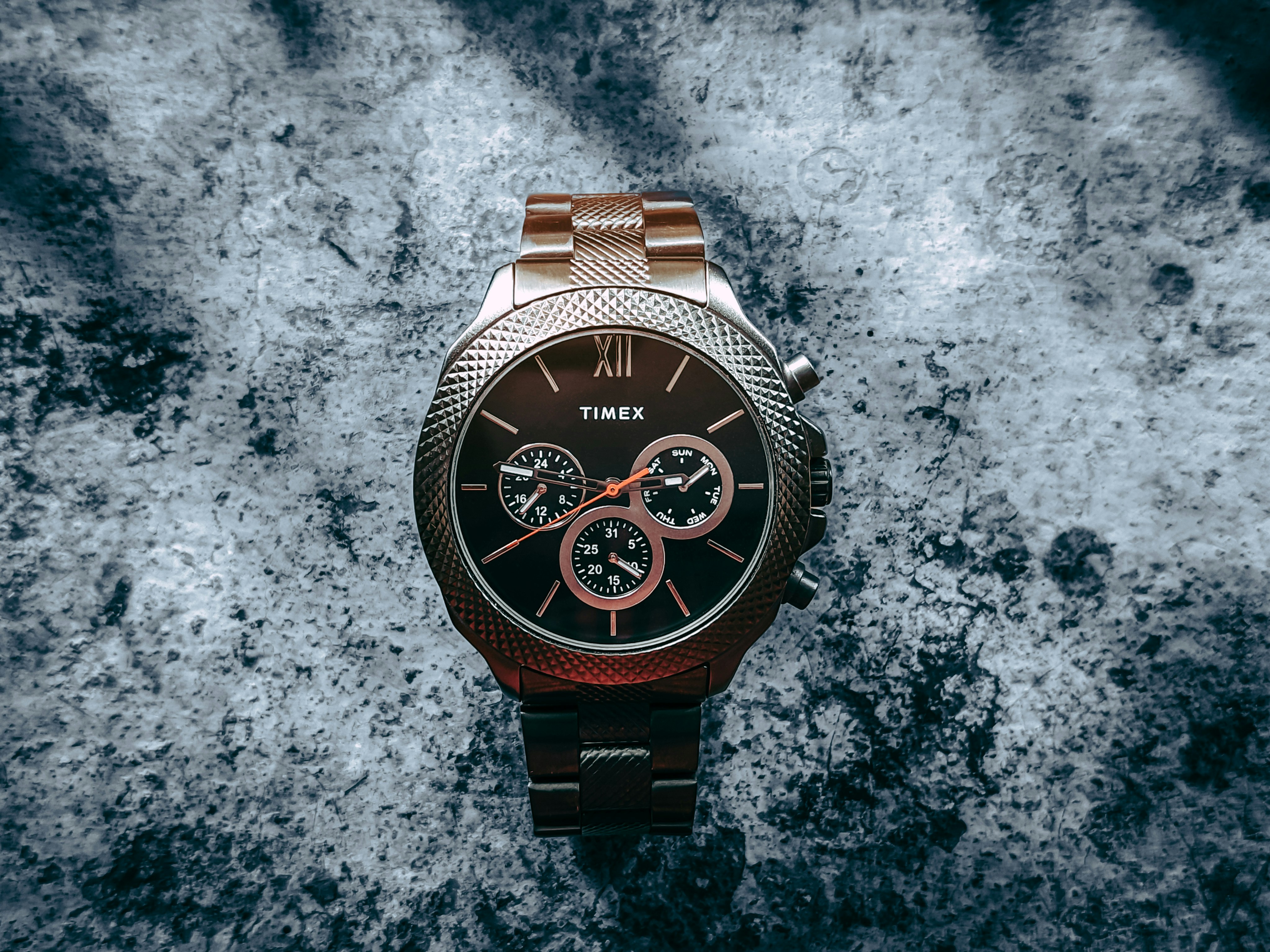 Close-up of a bronze Timex chronograph resting on a frost-speckled concrete surface. The metal bracelet gleams under soft light, highlighting texture and craftsmanship.