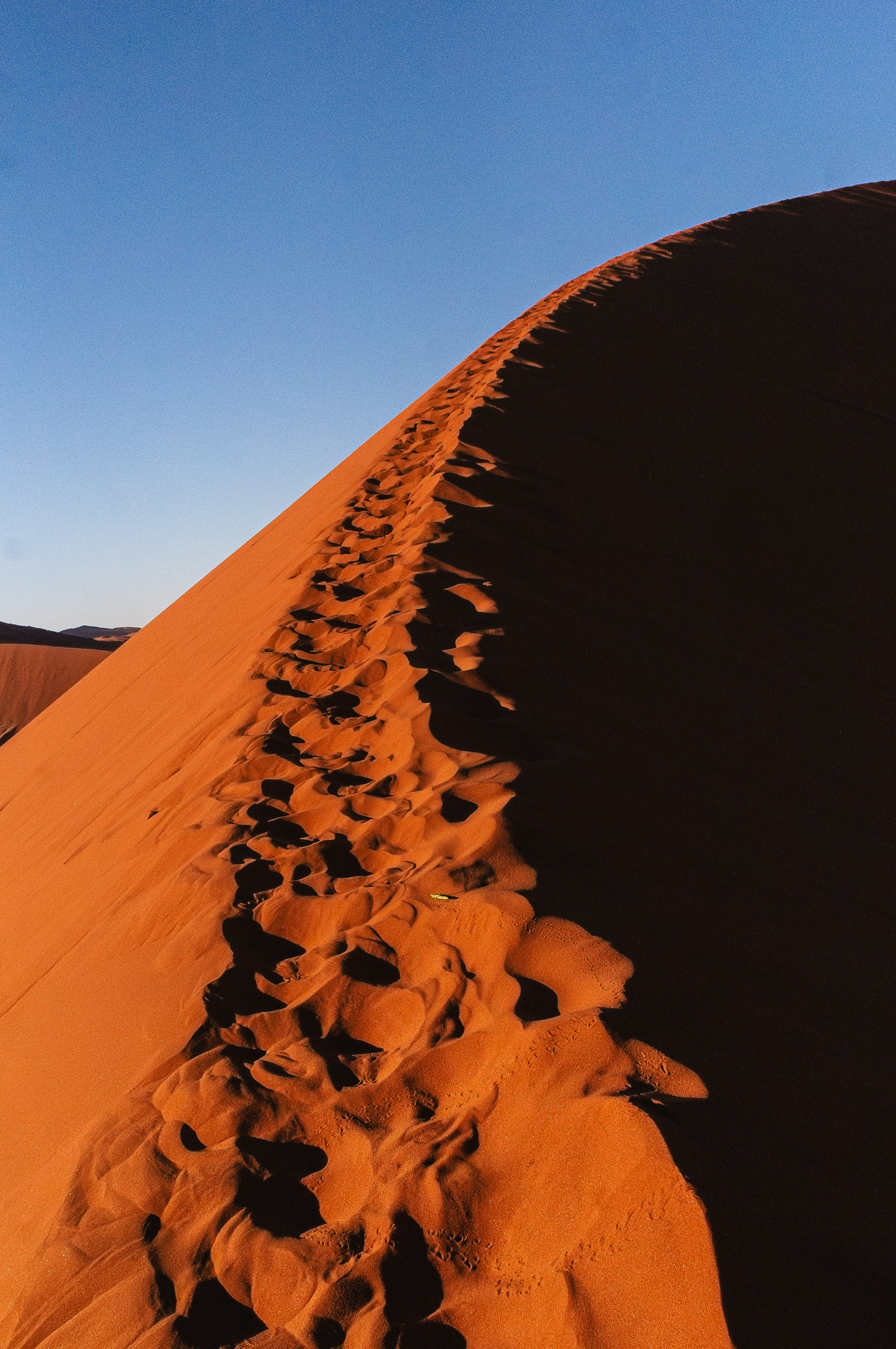 Crimson sand dunes glow under a clear blue sky, with footprints tracing a path along the dune crest.
