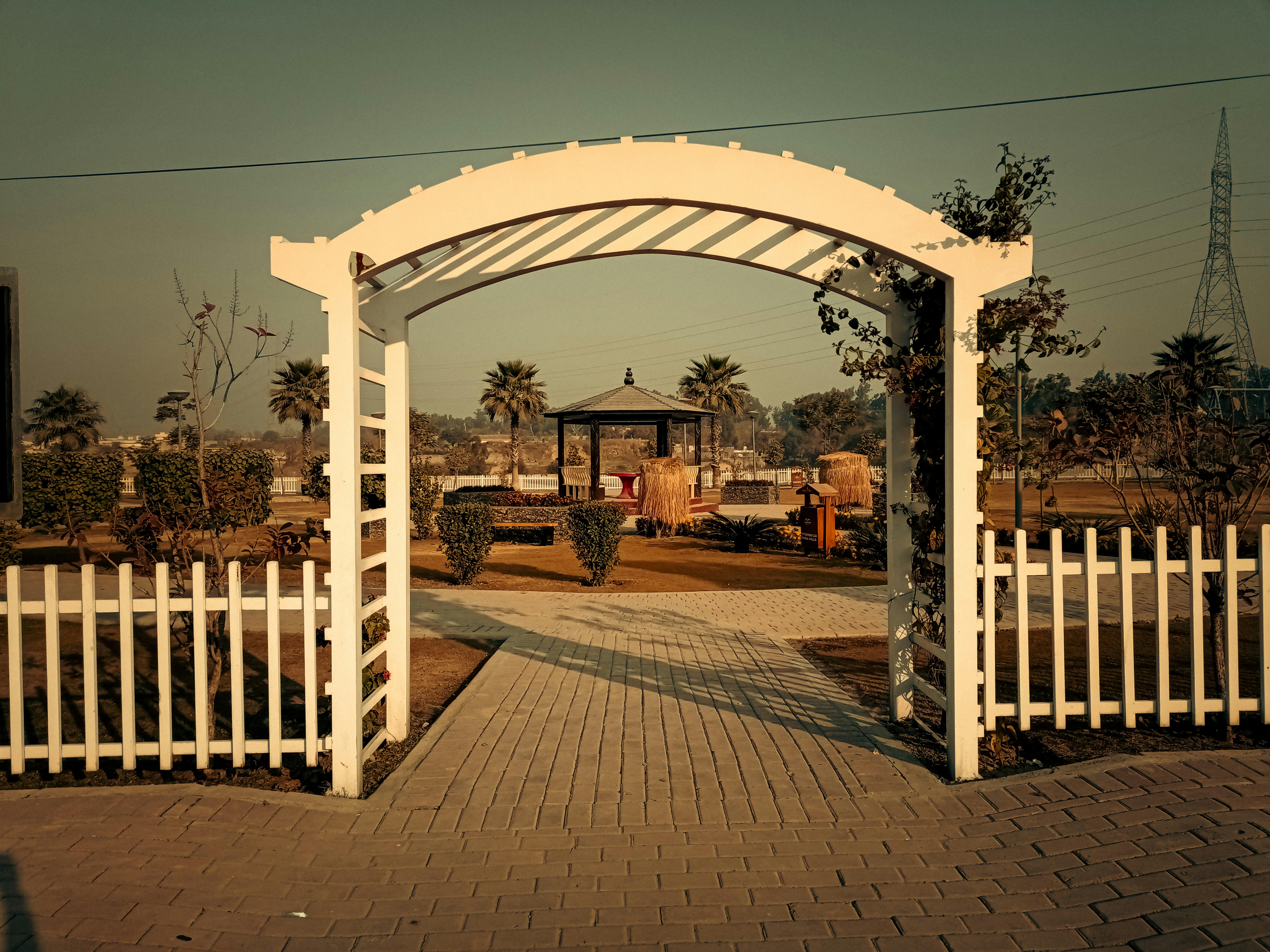 White archway leading into a serene park with a gazebo in the distance, framed by neatly trimmed hedges and palm trees.