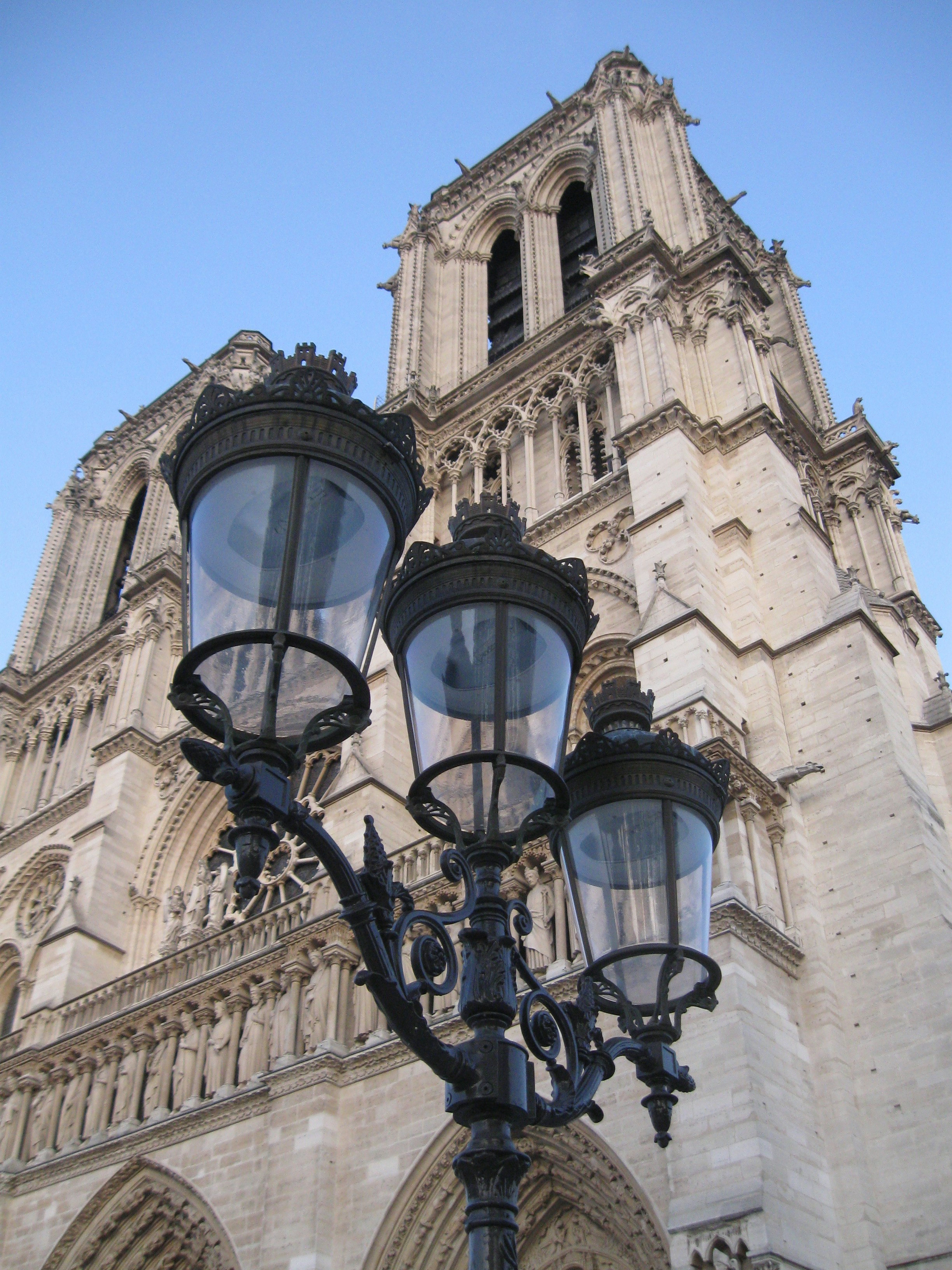 Photograph of a centuries-old Gothic cathedral rising behind an ornate iron street lamp cluster against a clear blue sky.