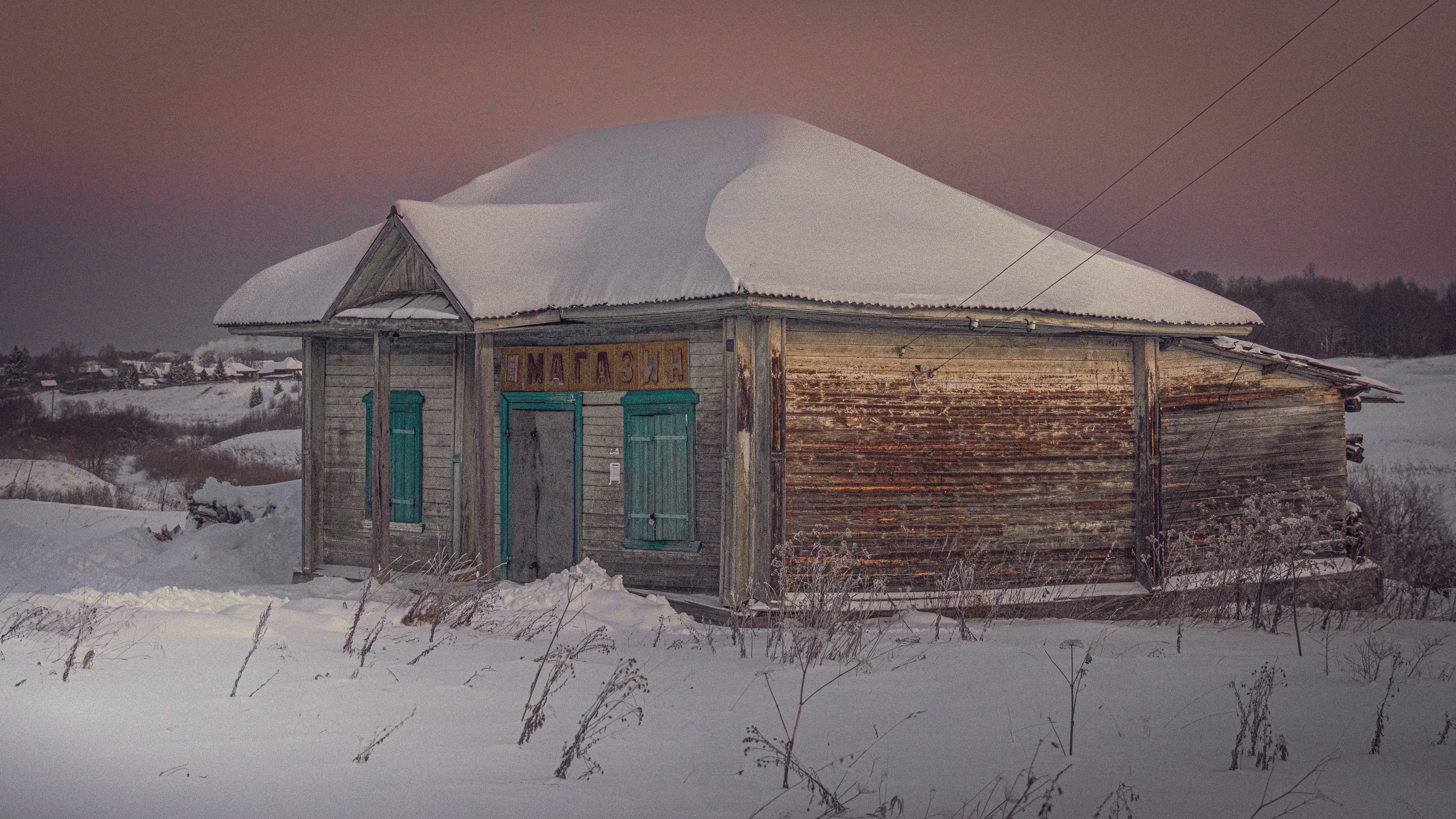 Snow-covered wooden cabin under a soft pink sky in a tranquil snowy landscape.