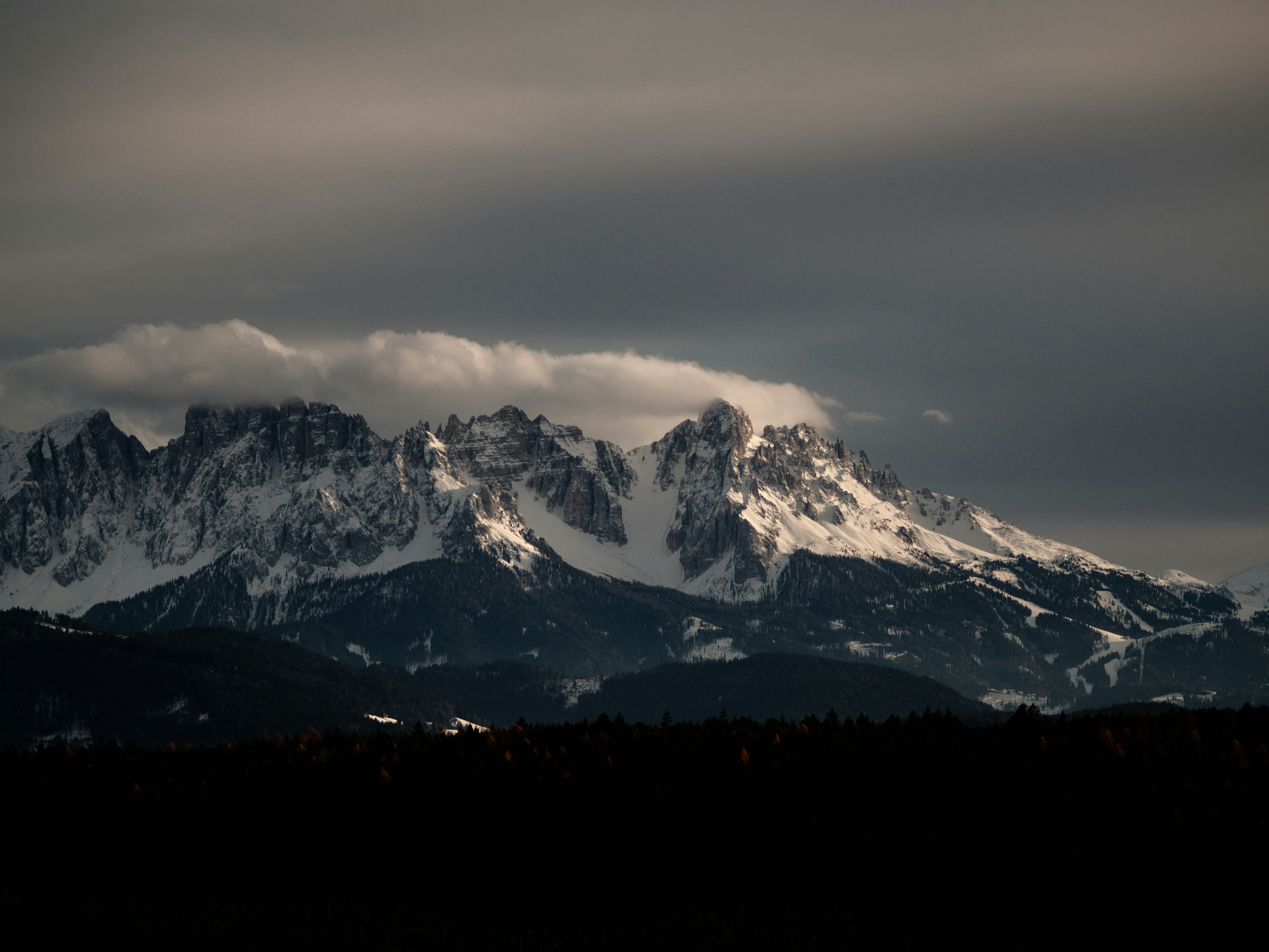 Snow-covered mountain range under dramatic cloud-filled sky at dusk.