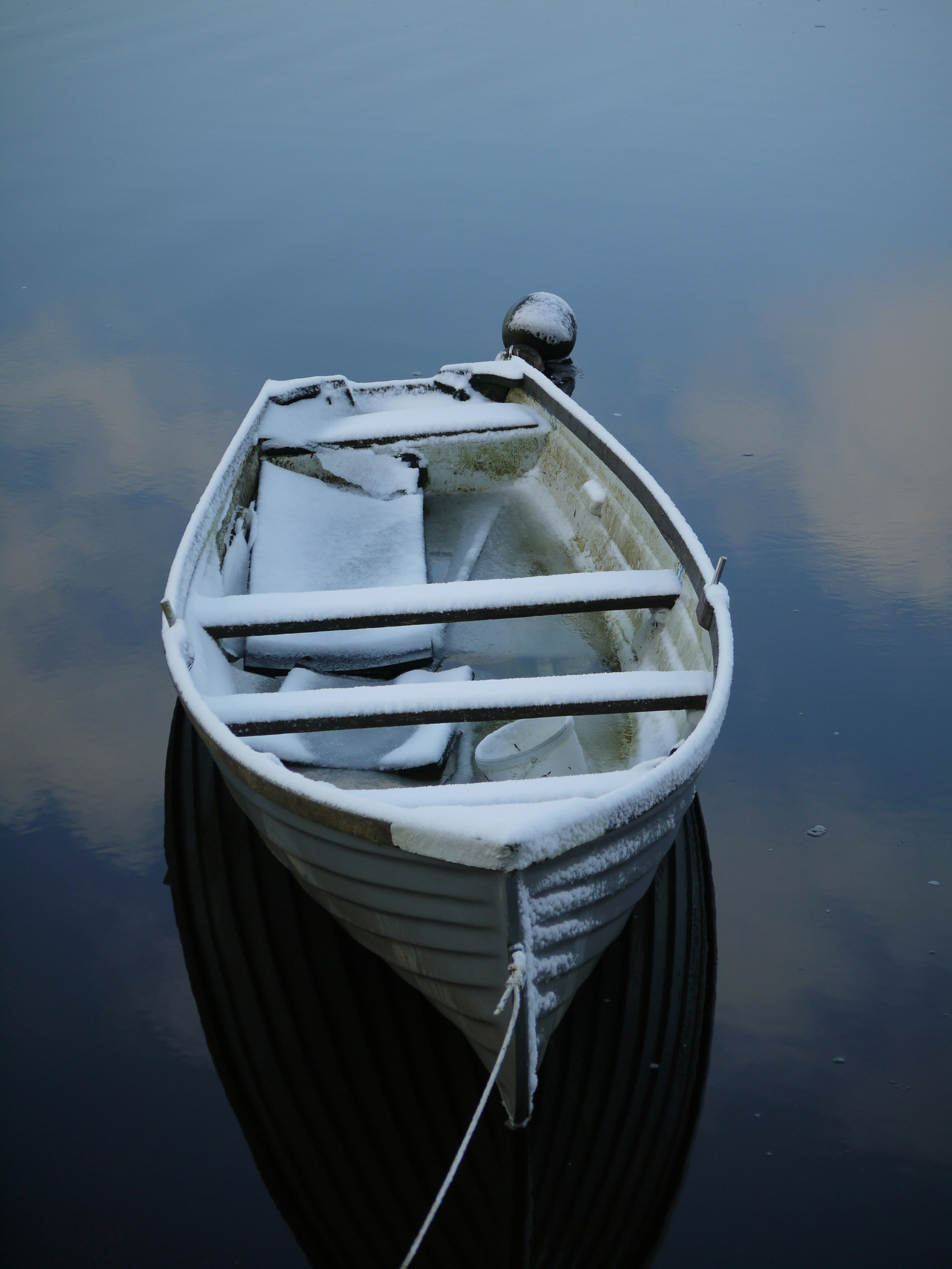 A small boat partially covered in snow floats peacefully on a calm body of water, reflecting the surrounding sky.