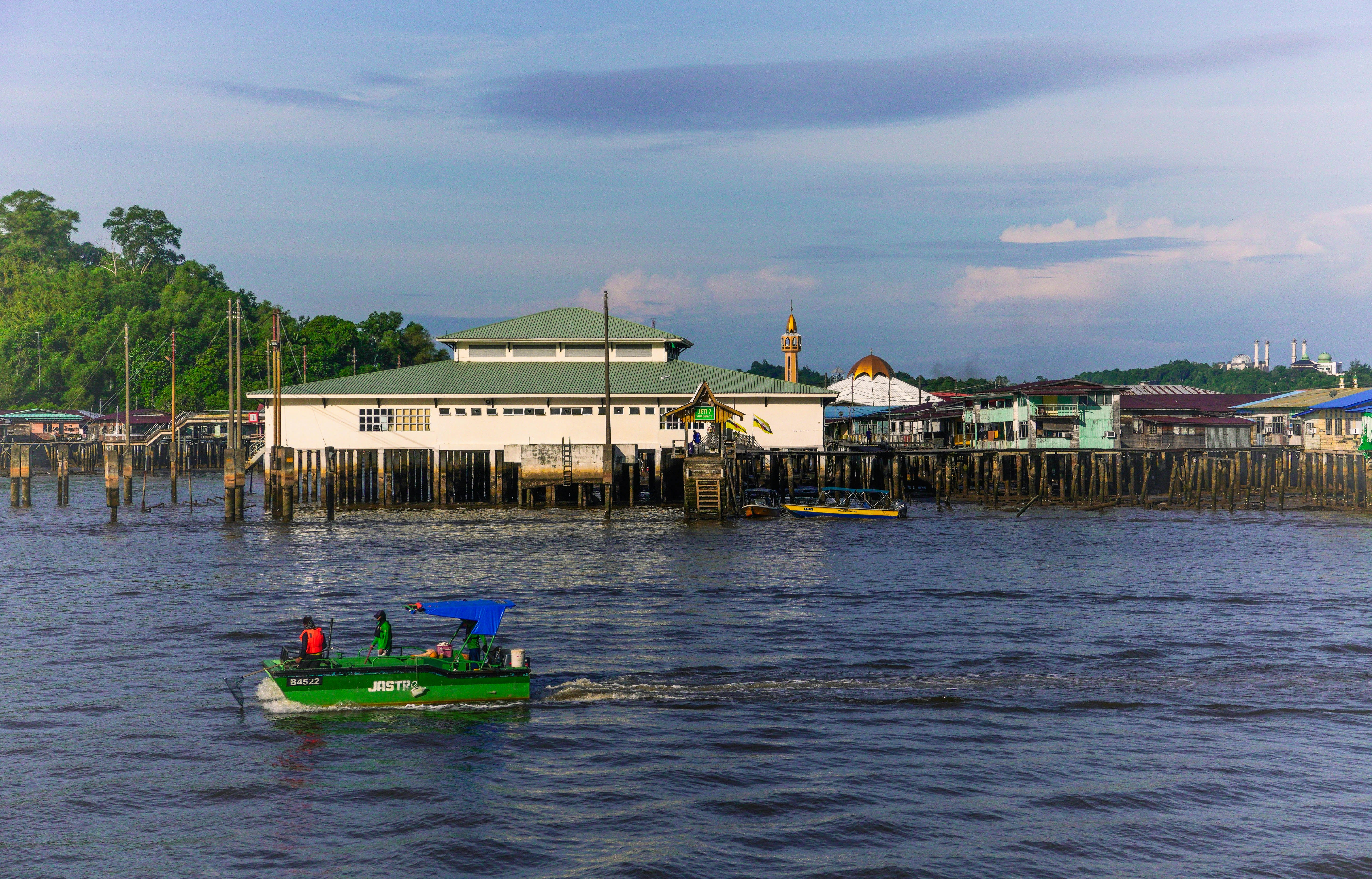 Green boat with blue canopy glides past wooden stilt houses on a calm river under a soft sky.