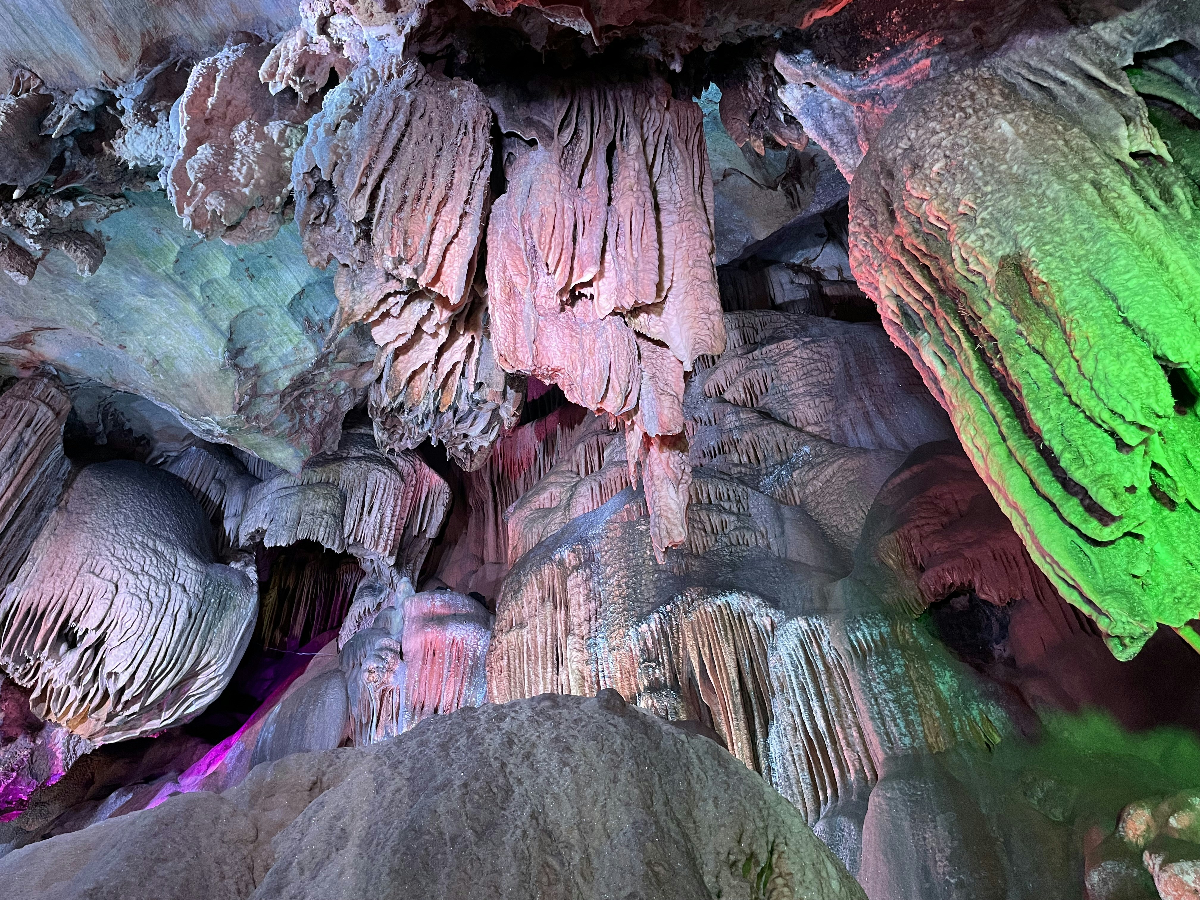 Stalactites and stalagmites in a cave illuminated by vibrant green and purple lights.