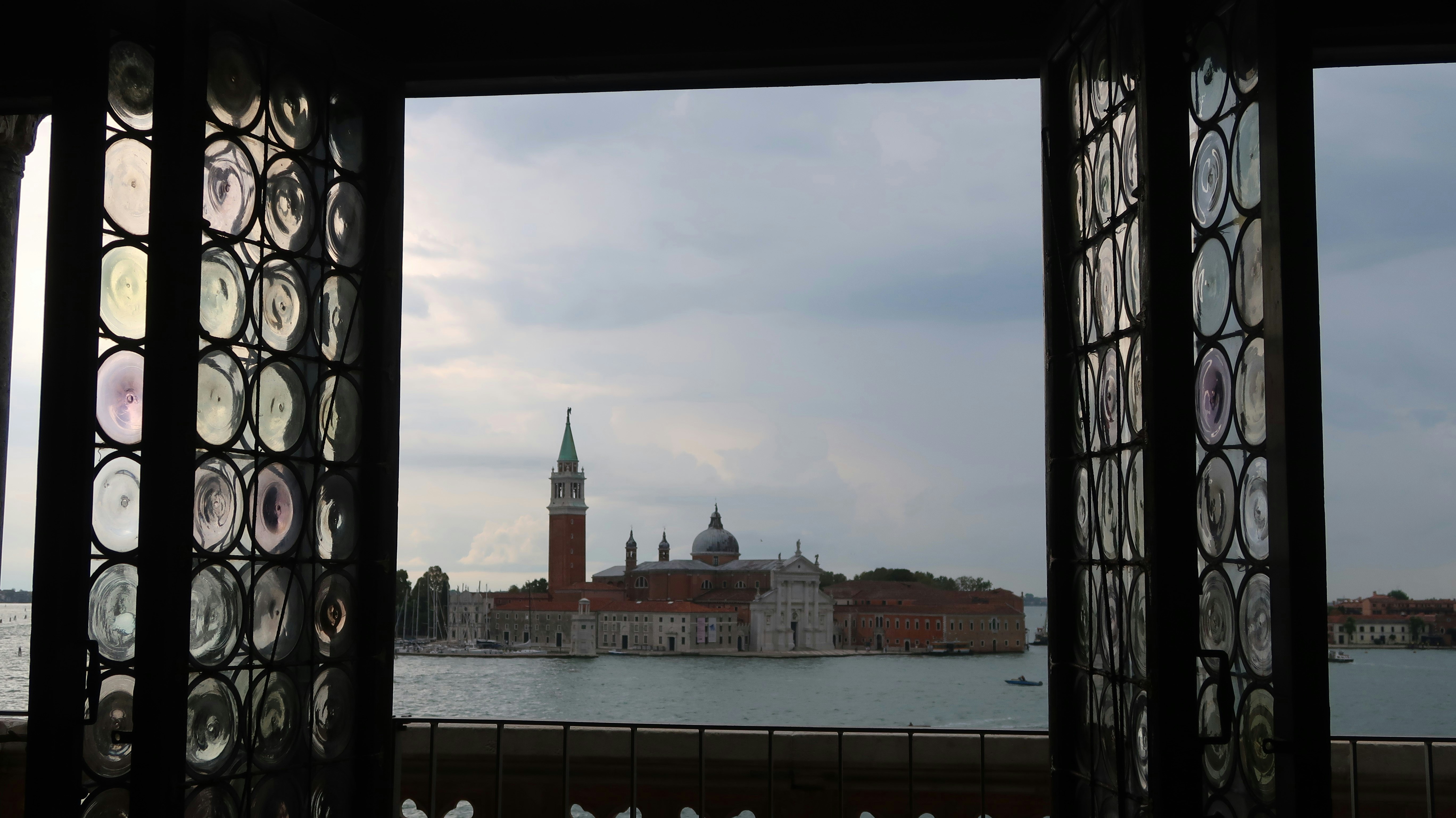 View of San Giorgio Maggiore island framed by ornate glass windows, showcasing the serene waters of Venice under a cloudy sky.