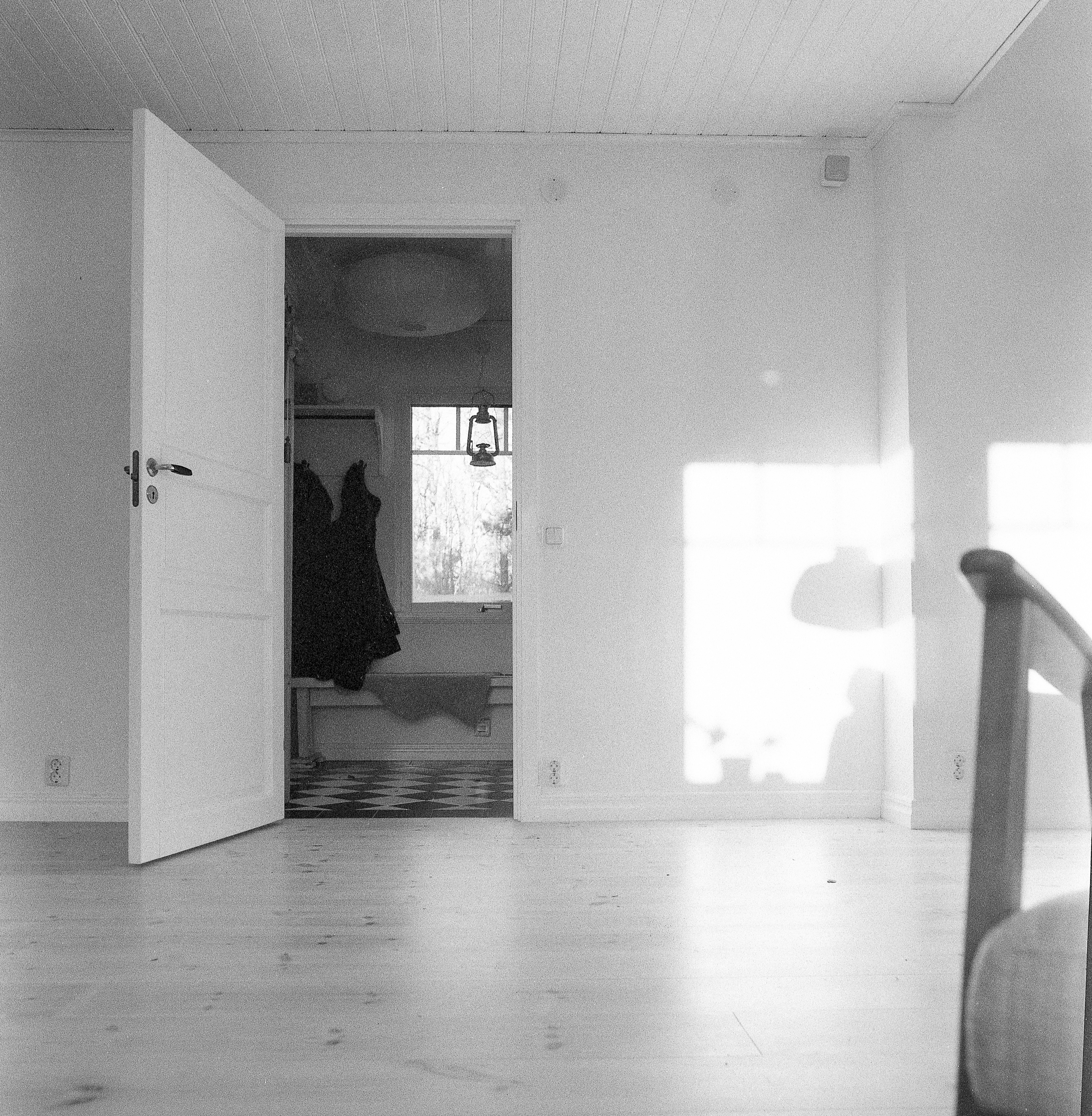 High-contrast black-and-white photograph of an empty living space with an open door leading to a second room with a checkered floor; sunlight creates bold geometric shadows.