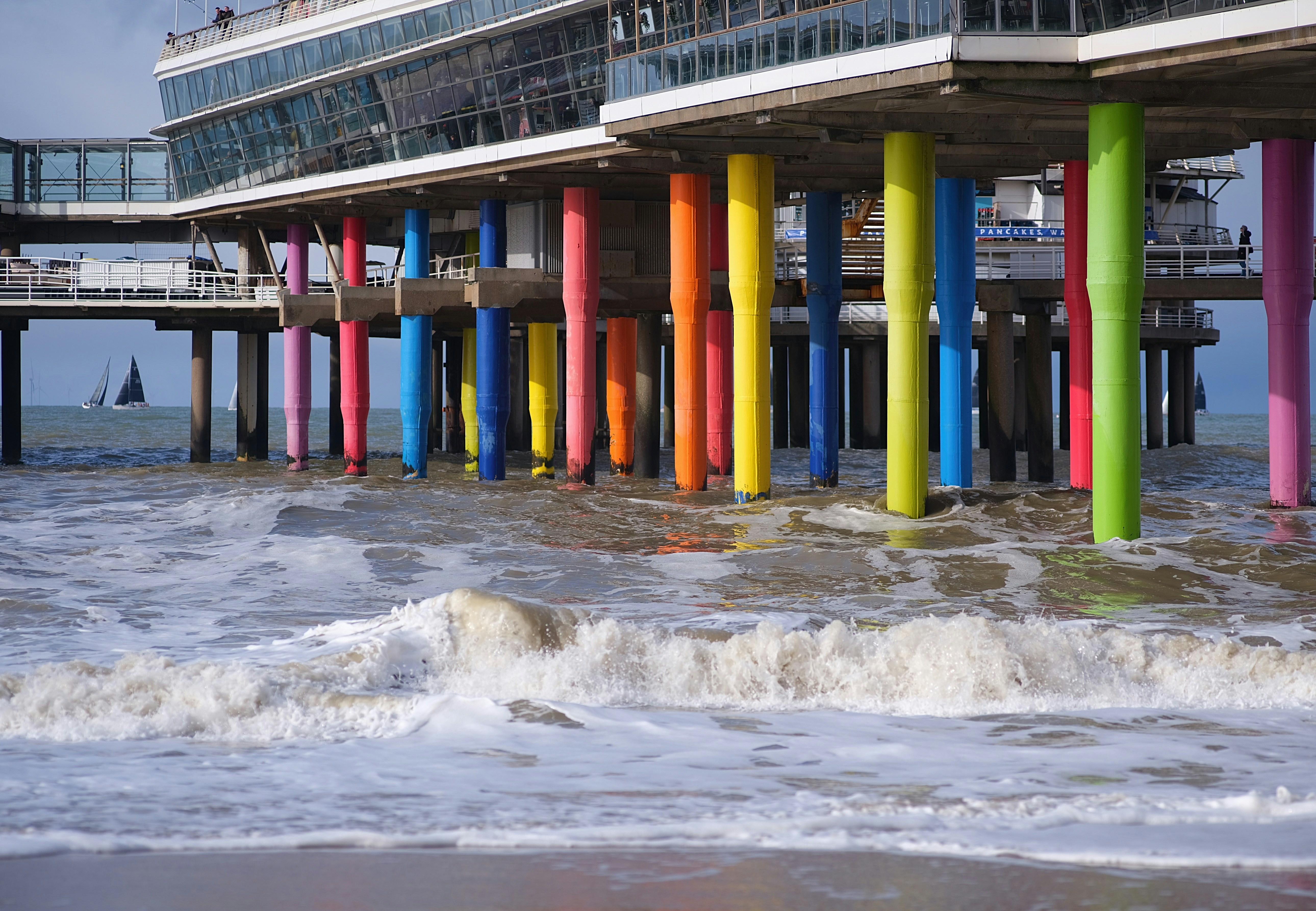 Rainbow Colonnade Under the Pier: Waves, Colorful Supports, and a Distant Sailboat