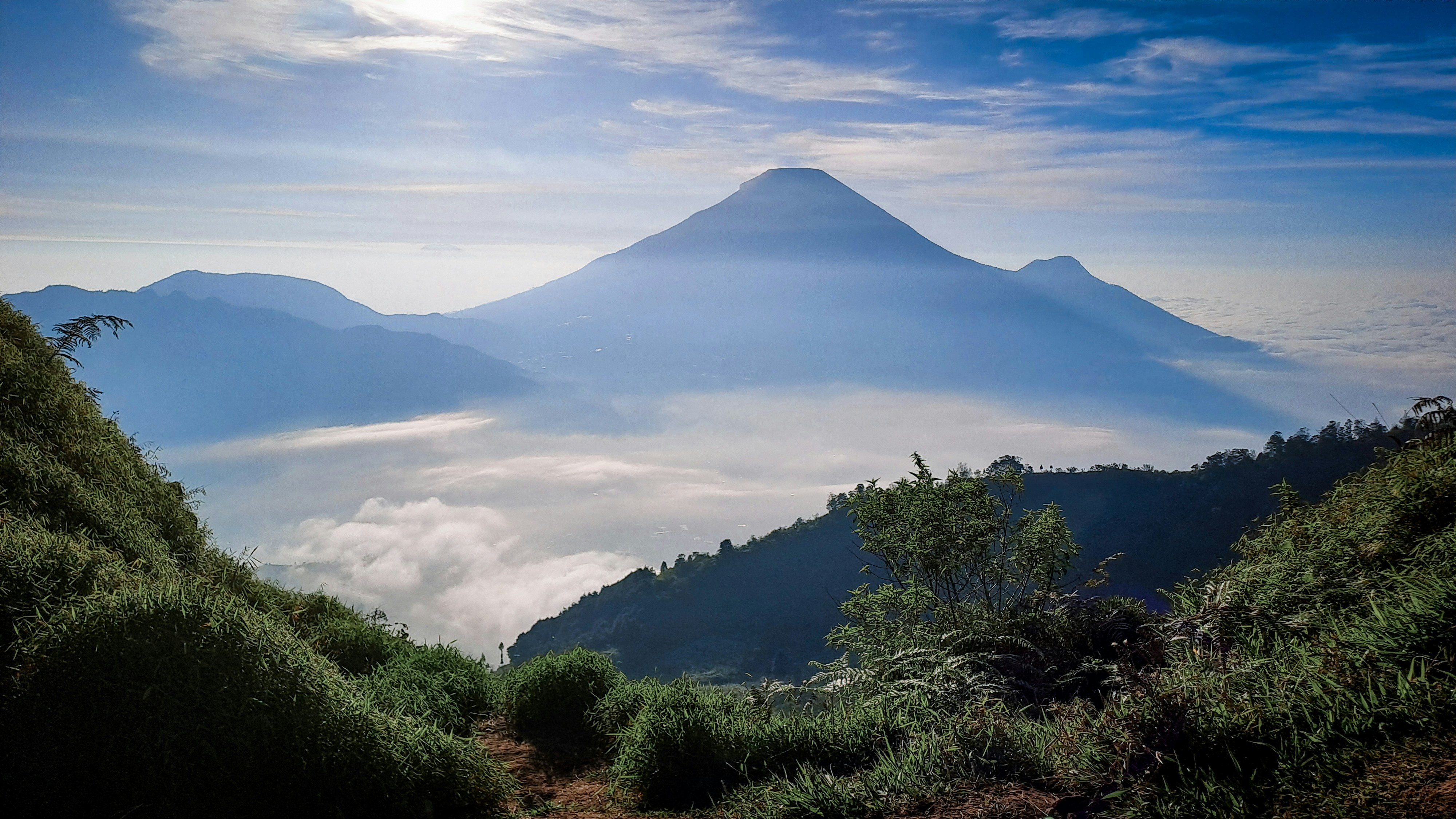 Lush green hills lead to a distant volcano shrouded in mist, with clouds rolling beneath the peaks. The serene landscape captures the tranquility of nature's grandeur.