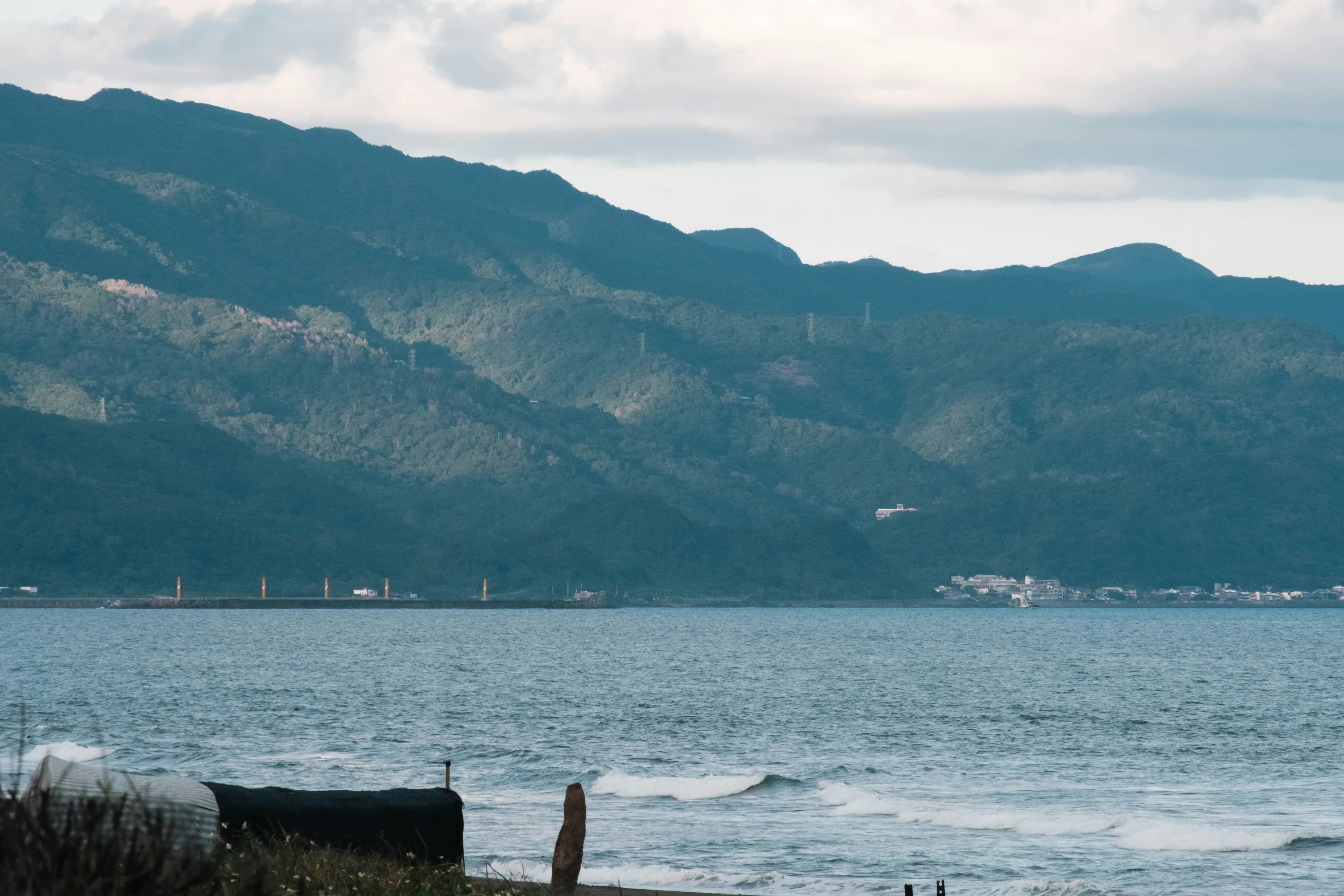 Waves gently lap against the shore with lush green mountains providing a serene backdrop under a cloudy sky.