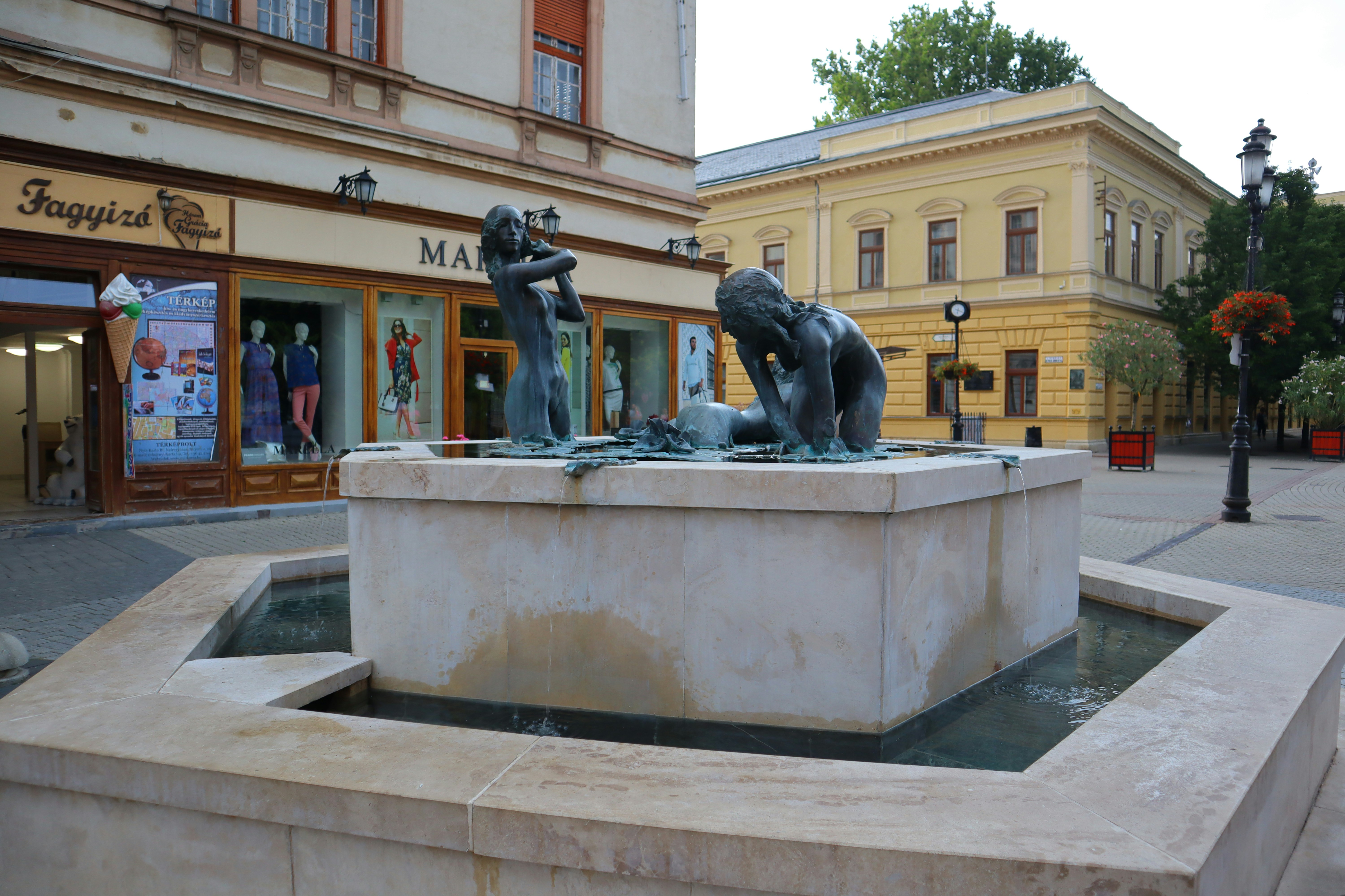 Bronze sculpture of figures around a fountain in a European square surrounded by historic buildings.
