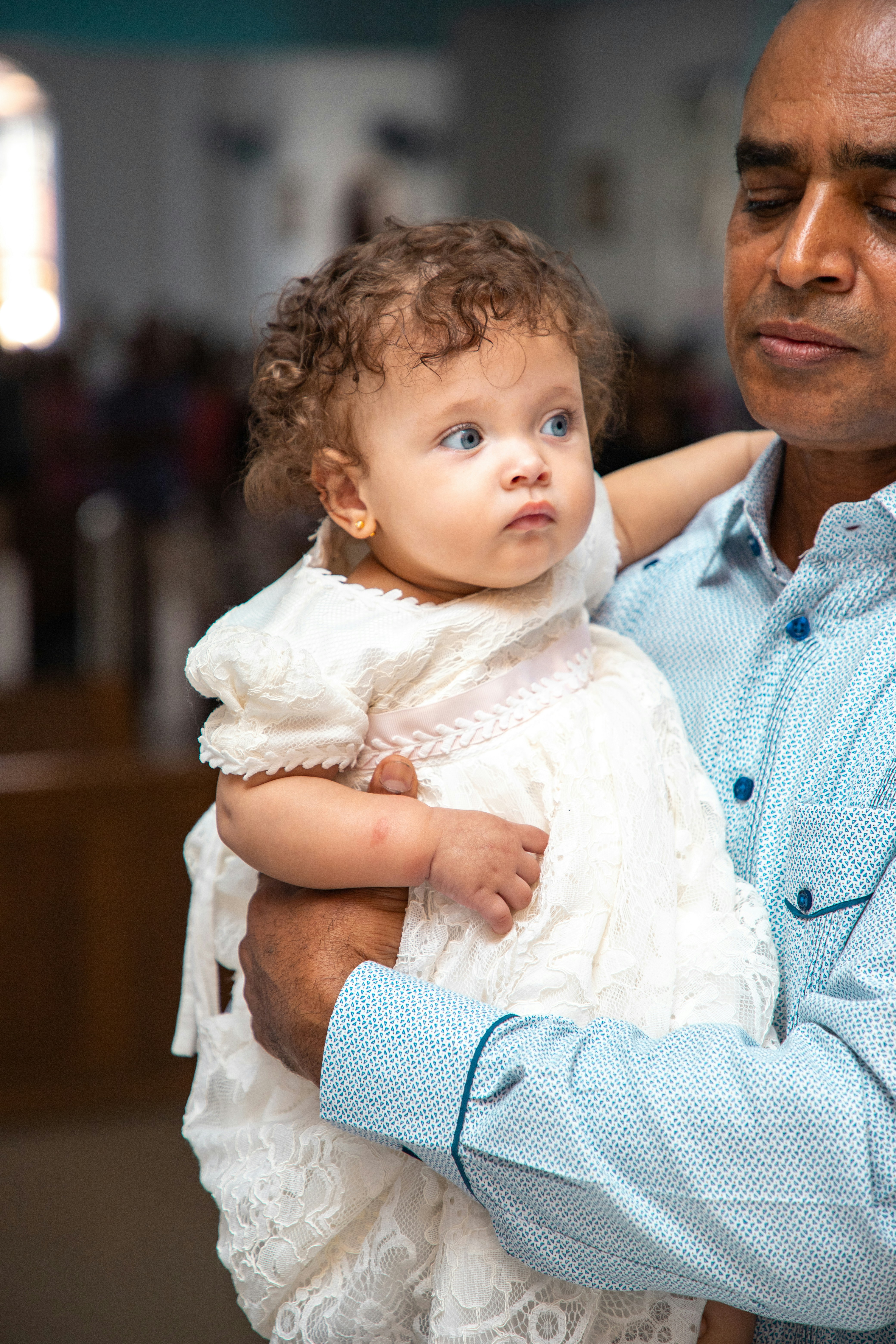 Child in lace dress held by an adult in a softly lit indoor setting.
