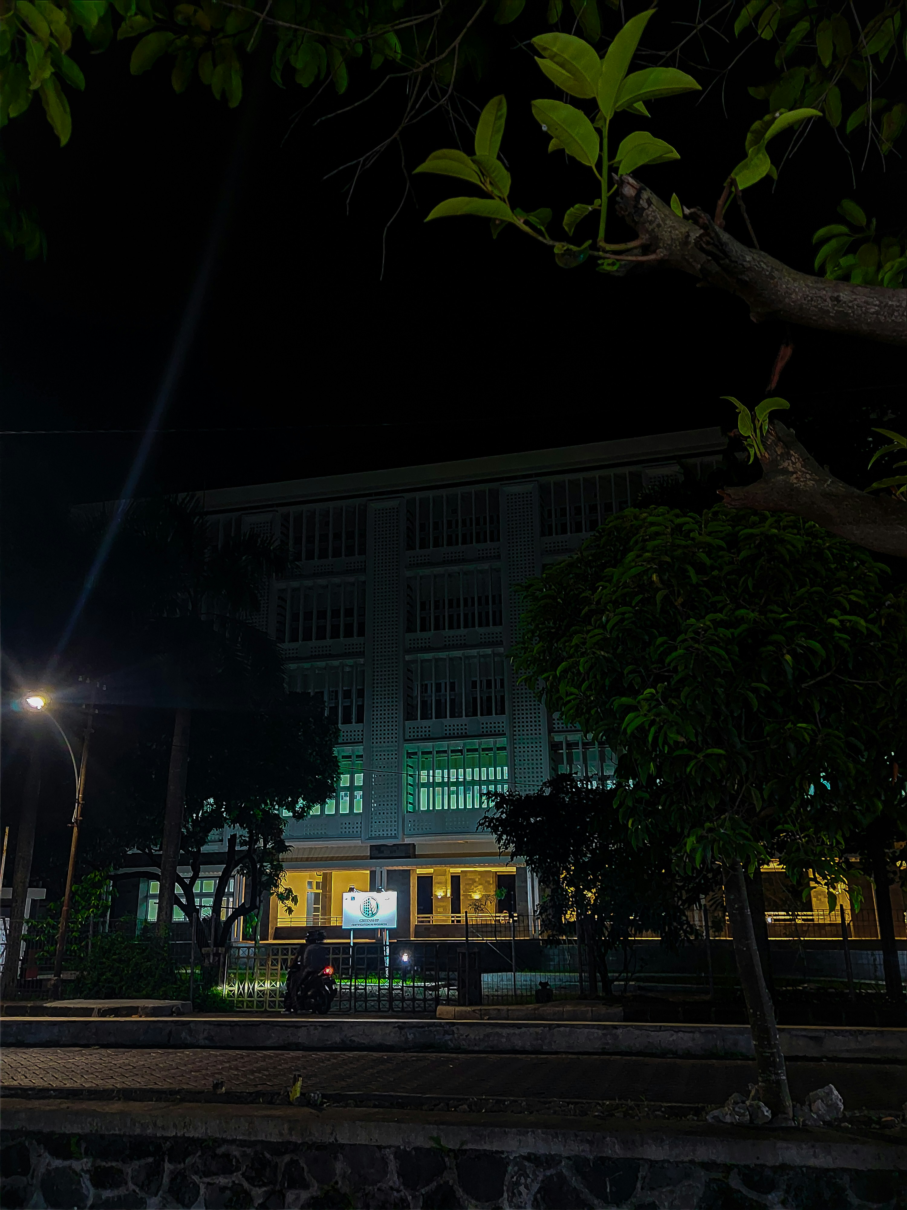 Nighttime campus facade photograph with a warmly lit entrance and visible signage. Trees frame the foreground against a dark sky.