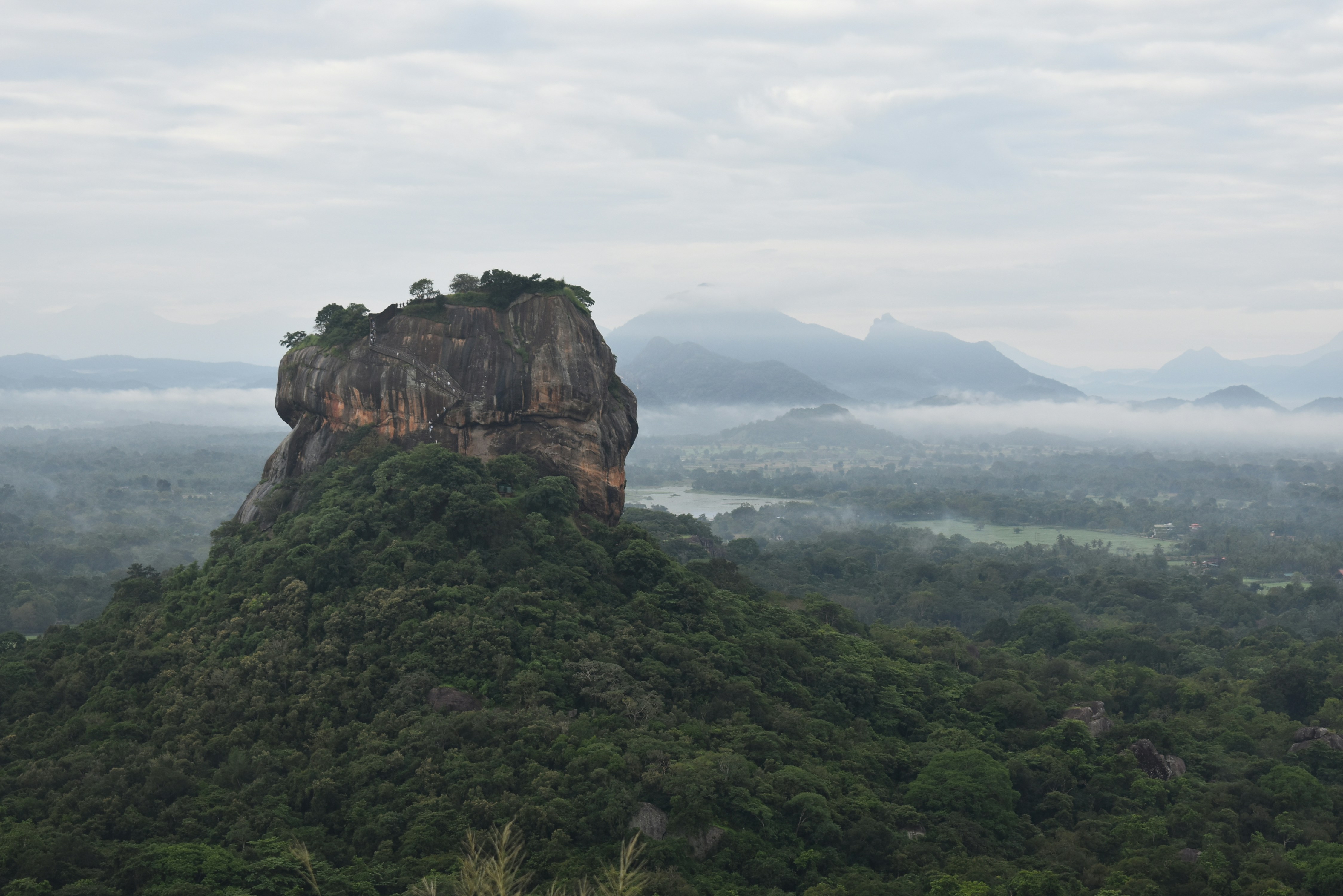 Prominent rock formation rises above lush greenery, shrouded in mist and framed by distant mountains. A serene landscape showcases the harmony between earth and sky.
