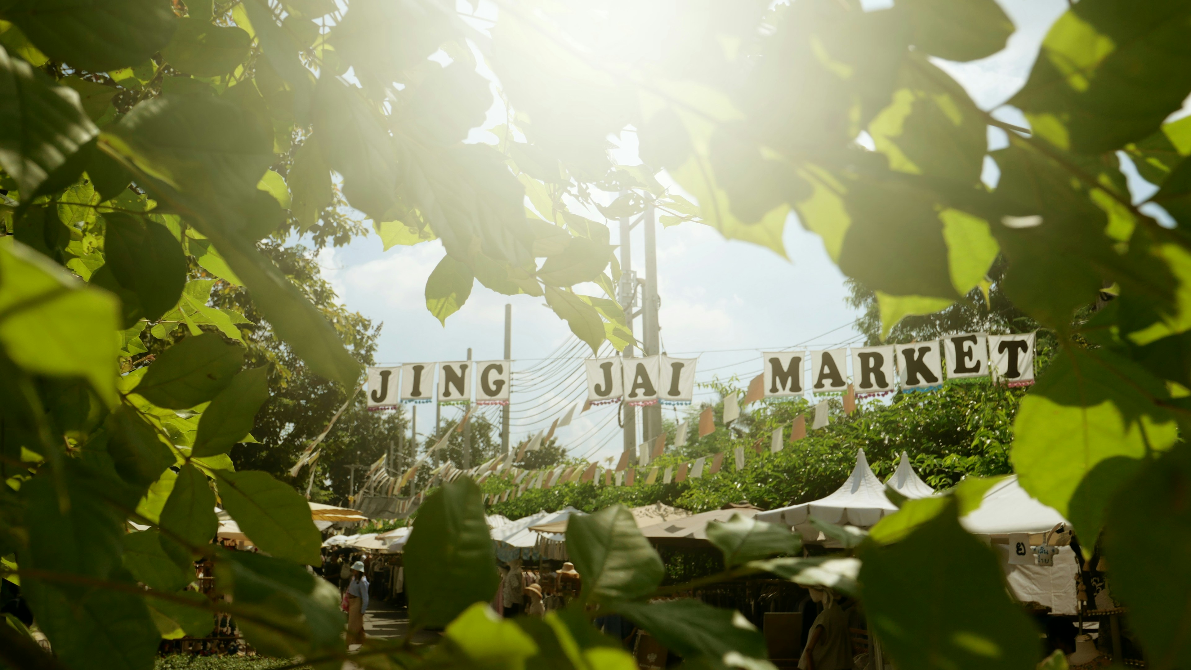 Photograph of the Jing Jai Market sign framed by a bright green canopy. Market stalls extend beneath in daylight.