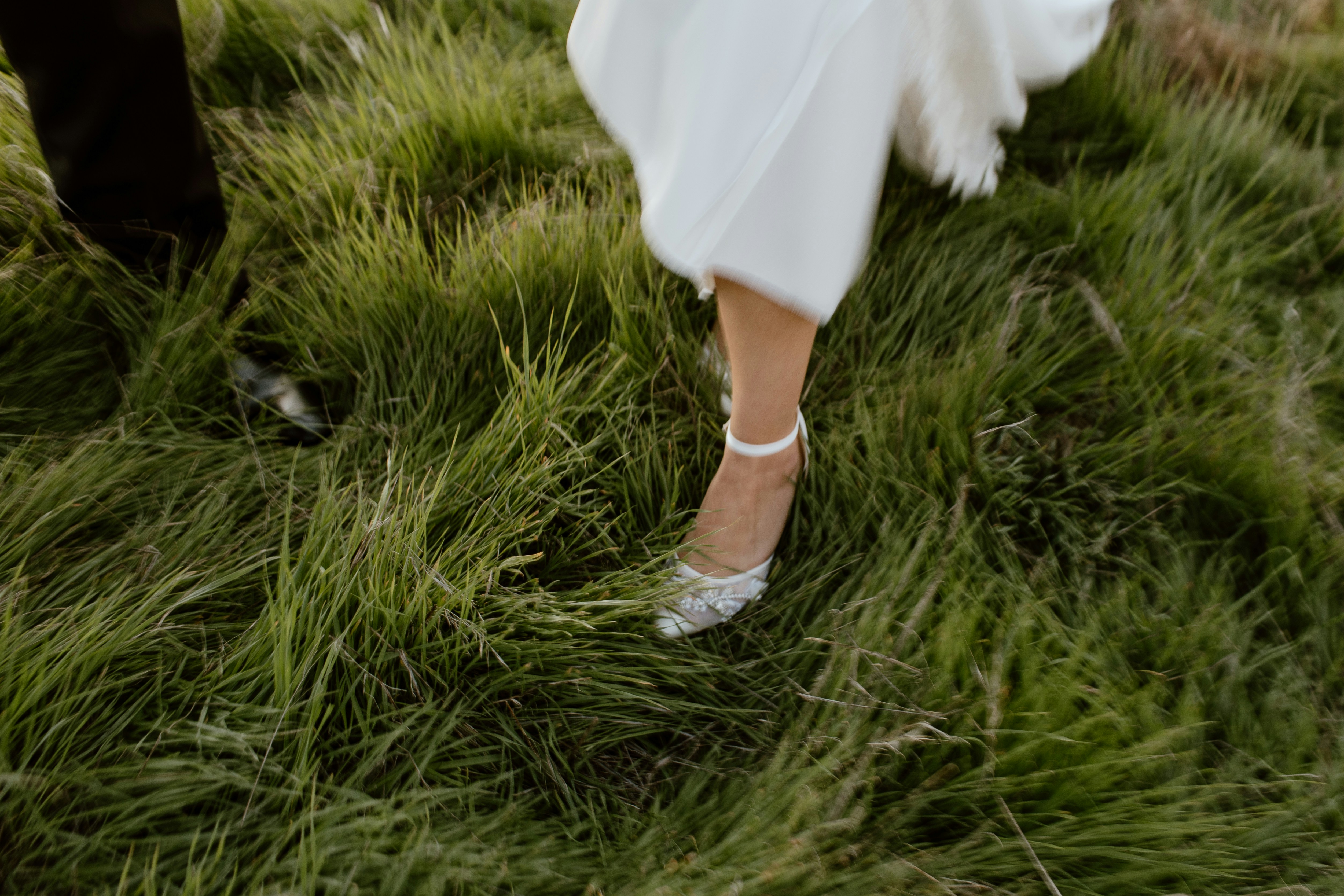 A close-up photograph of a white satin wedding shoe stepping through tussled grass, with a flowing white dress visible. The frame captures a candid, intimate moment in a natural setting.