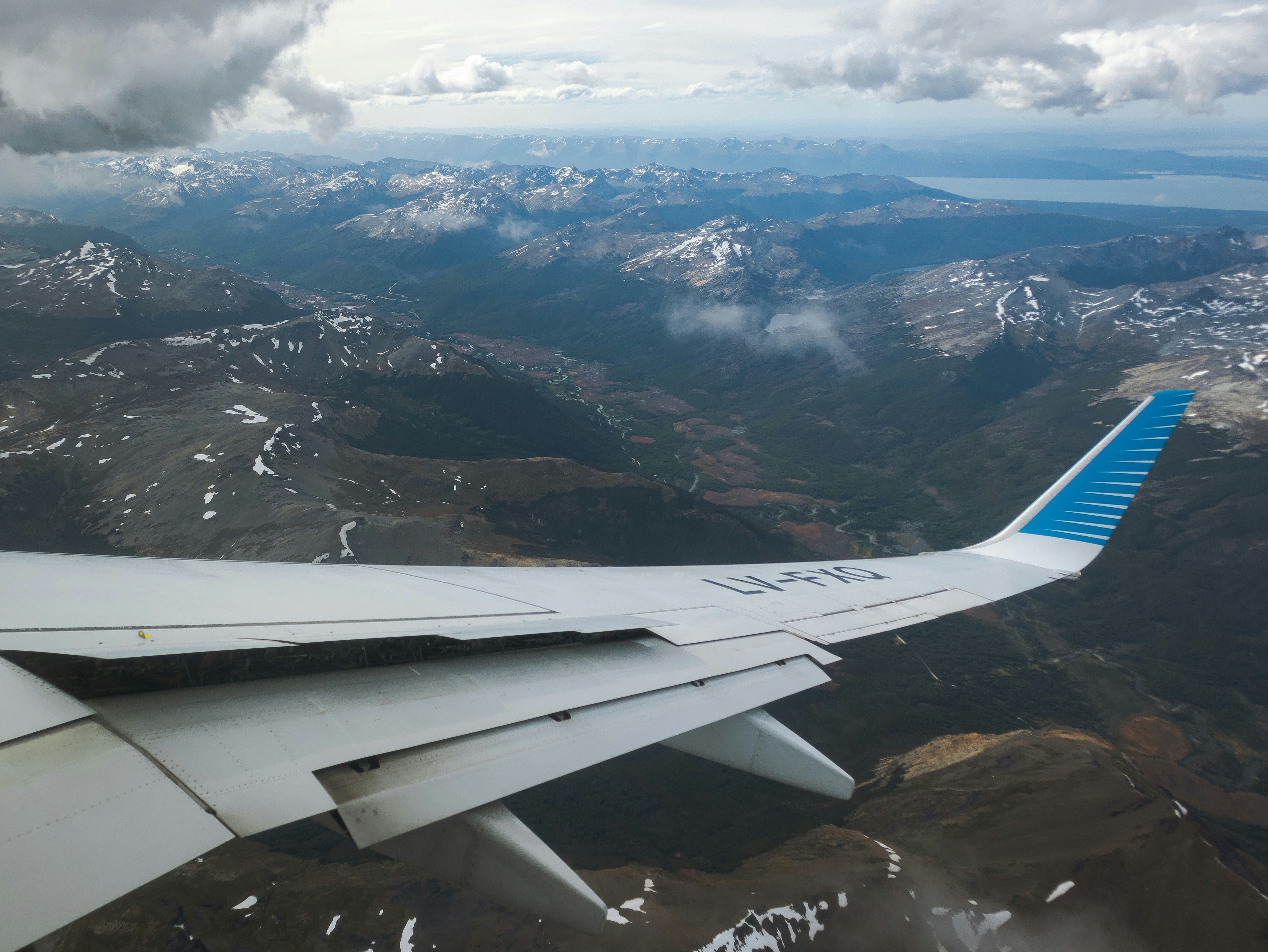 Airplane wing soaring above rugged, snow-capped mountains under a cloudy sky.