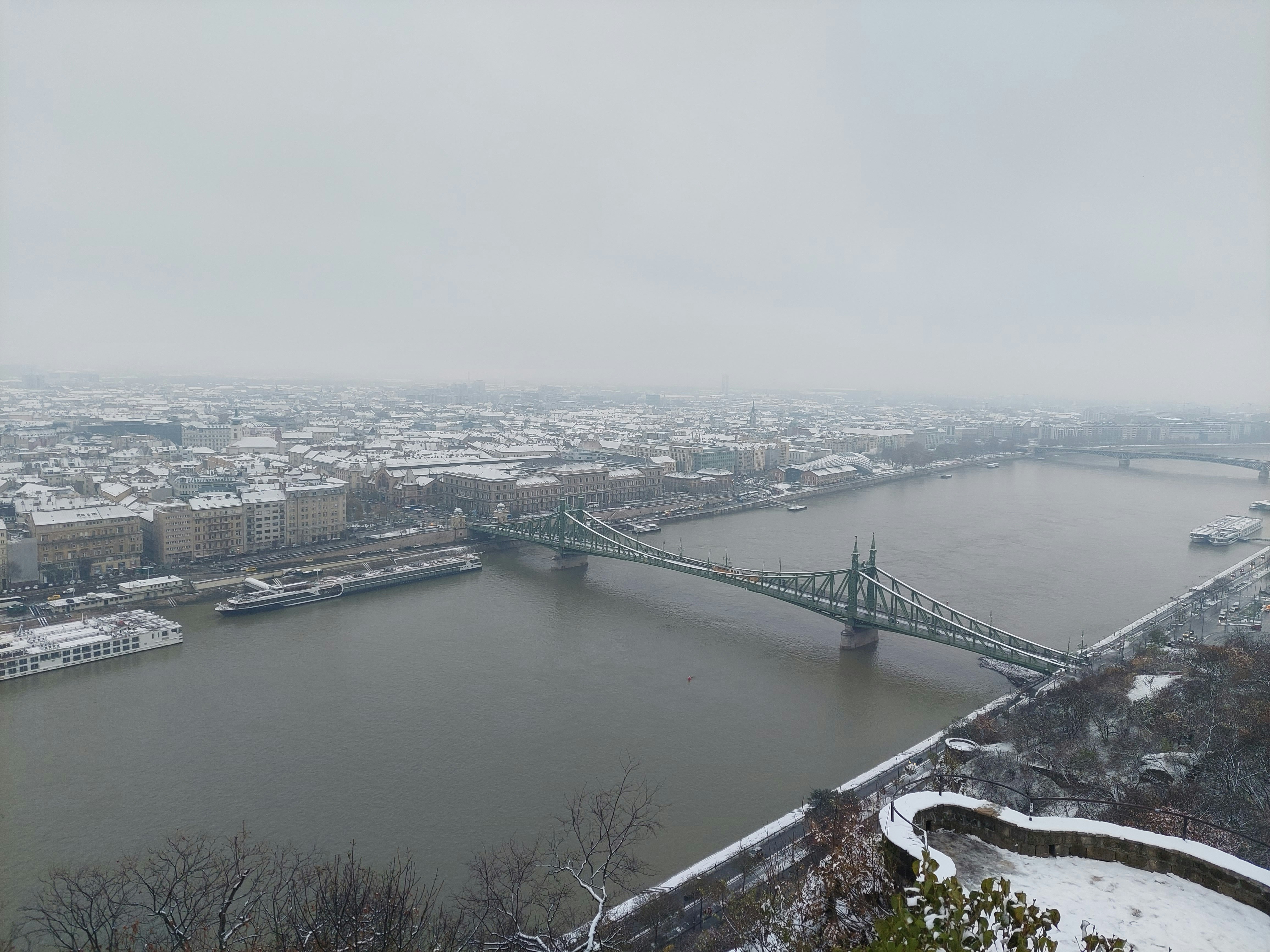 Overcast cityscape photograph of a steel suspension bridge spanning a wide river, with snow on the banks and rooftops in muted winter tones.