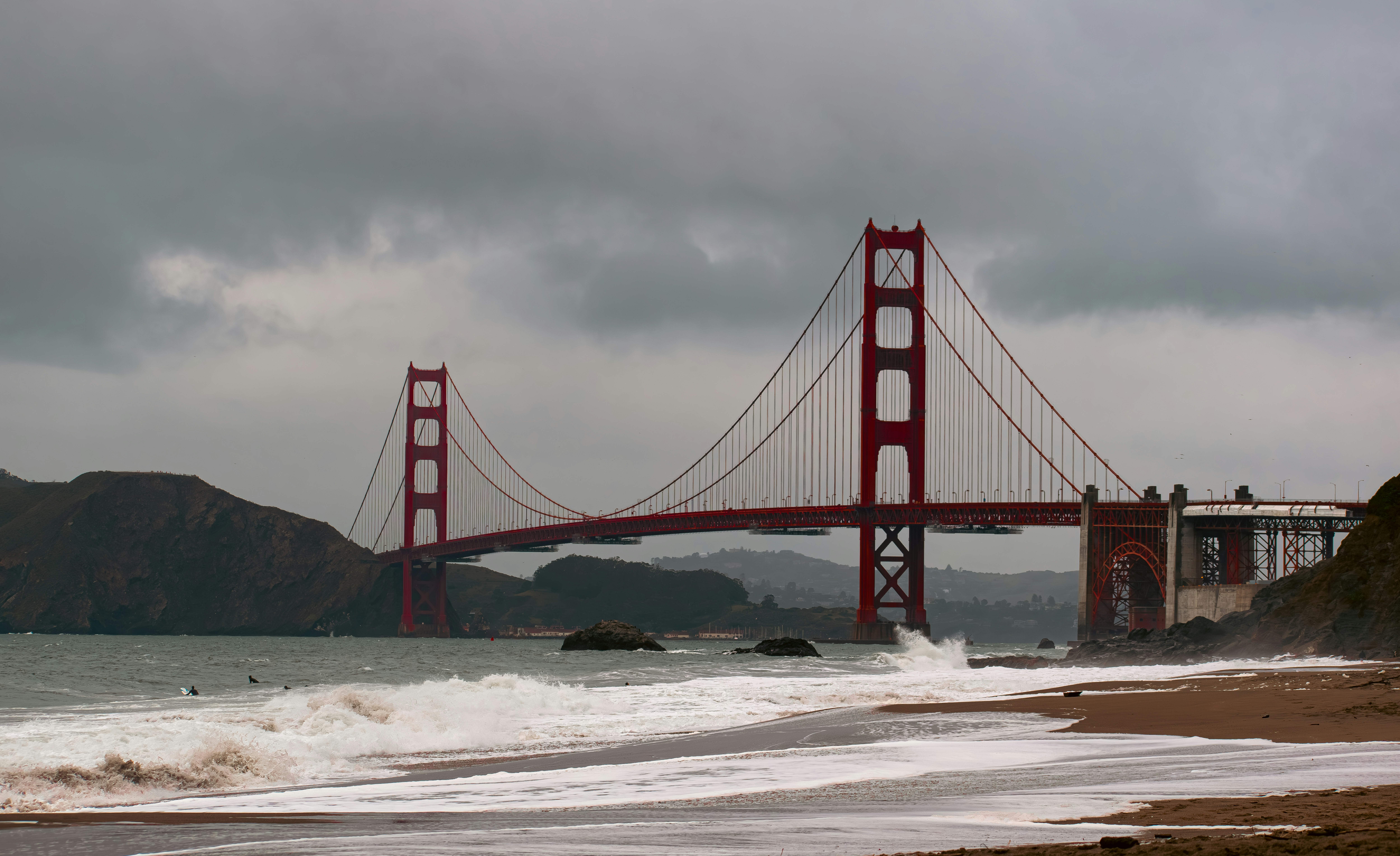Golden Gate Bridge under overcast skies with waves crashing on a sandy beach.