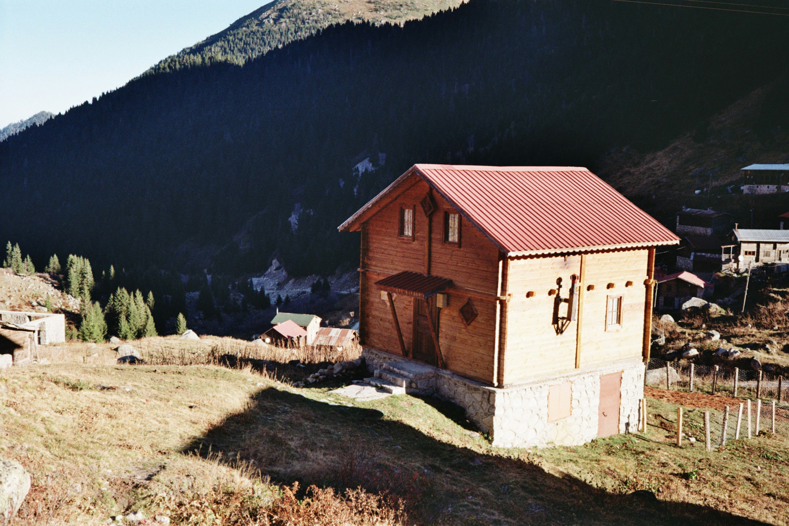 Photograph of a wooden alpine cabin set on a sunlit hillside with pine-covered mountains in the background.