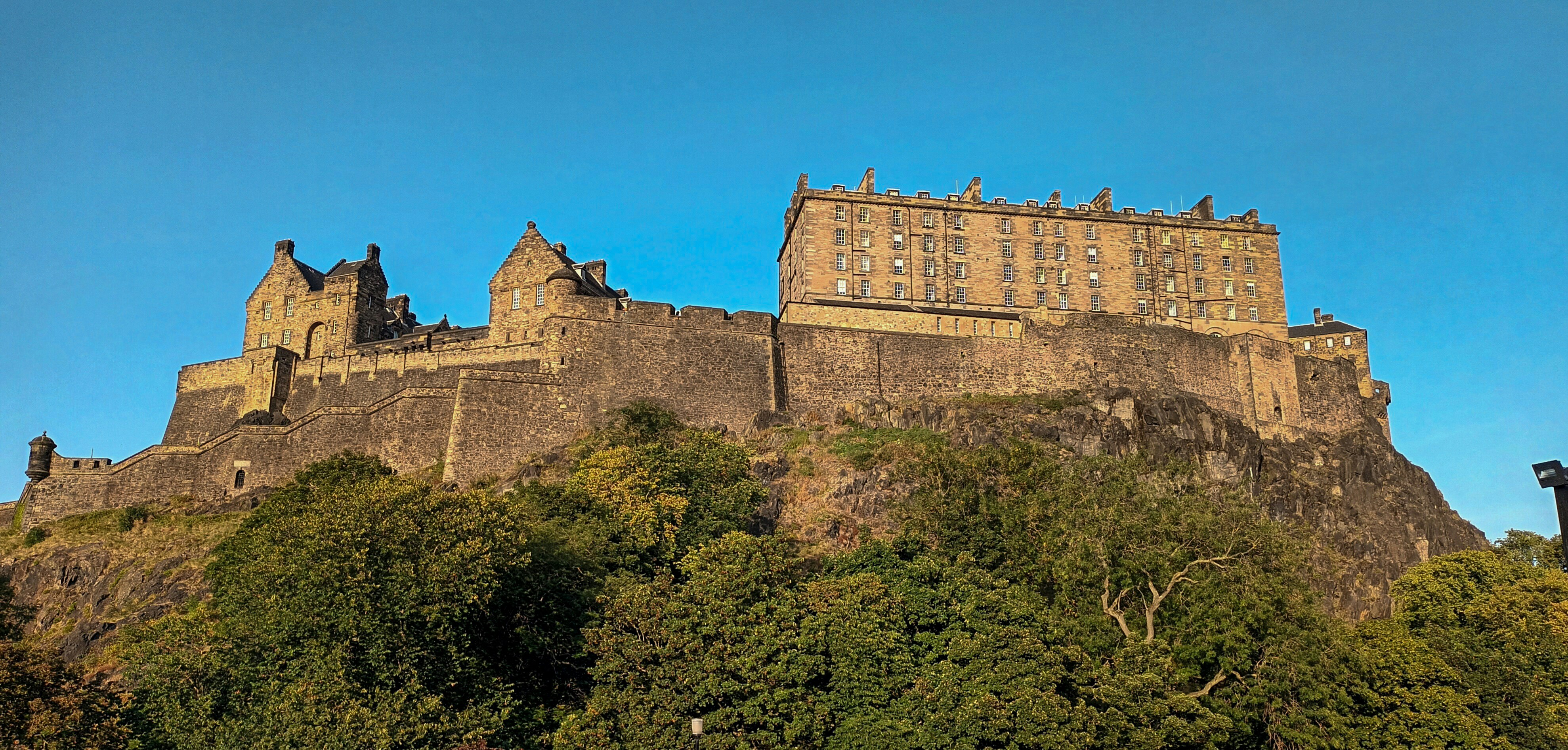 Stone hilltop fortress rises above a lush green canopy beneath a clear blue sky.