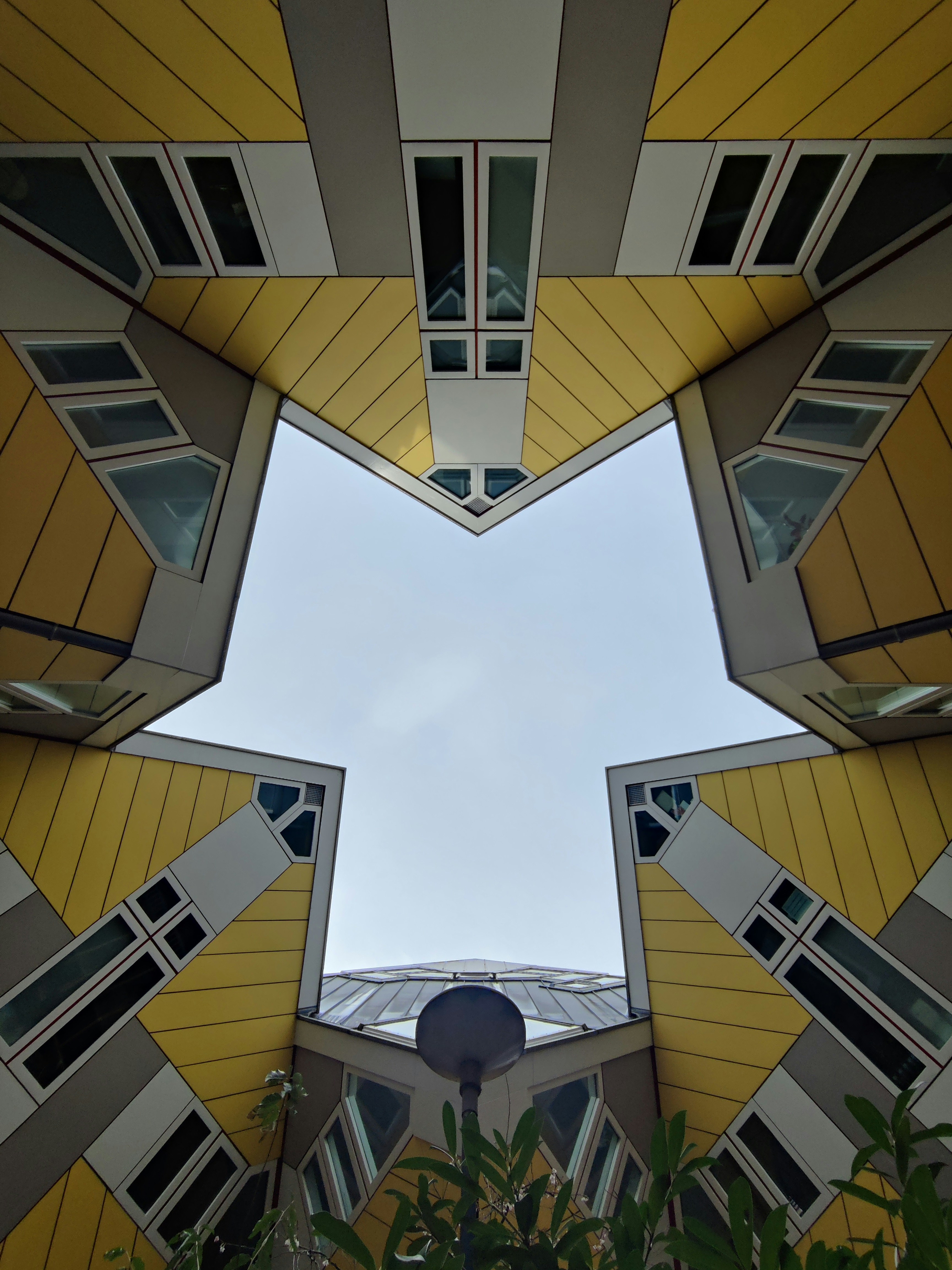 Photograph of yellow geometric buildings converging around a central blue sky, shot from a low angle between the structures. The composition highlights converging lines and negative space.