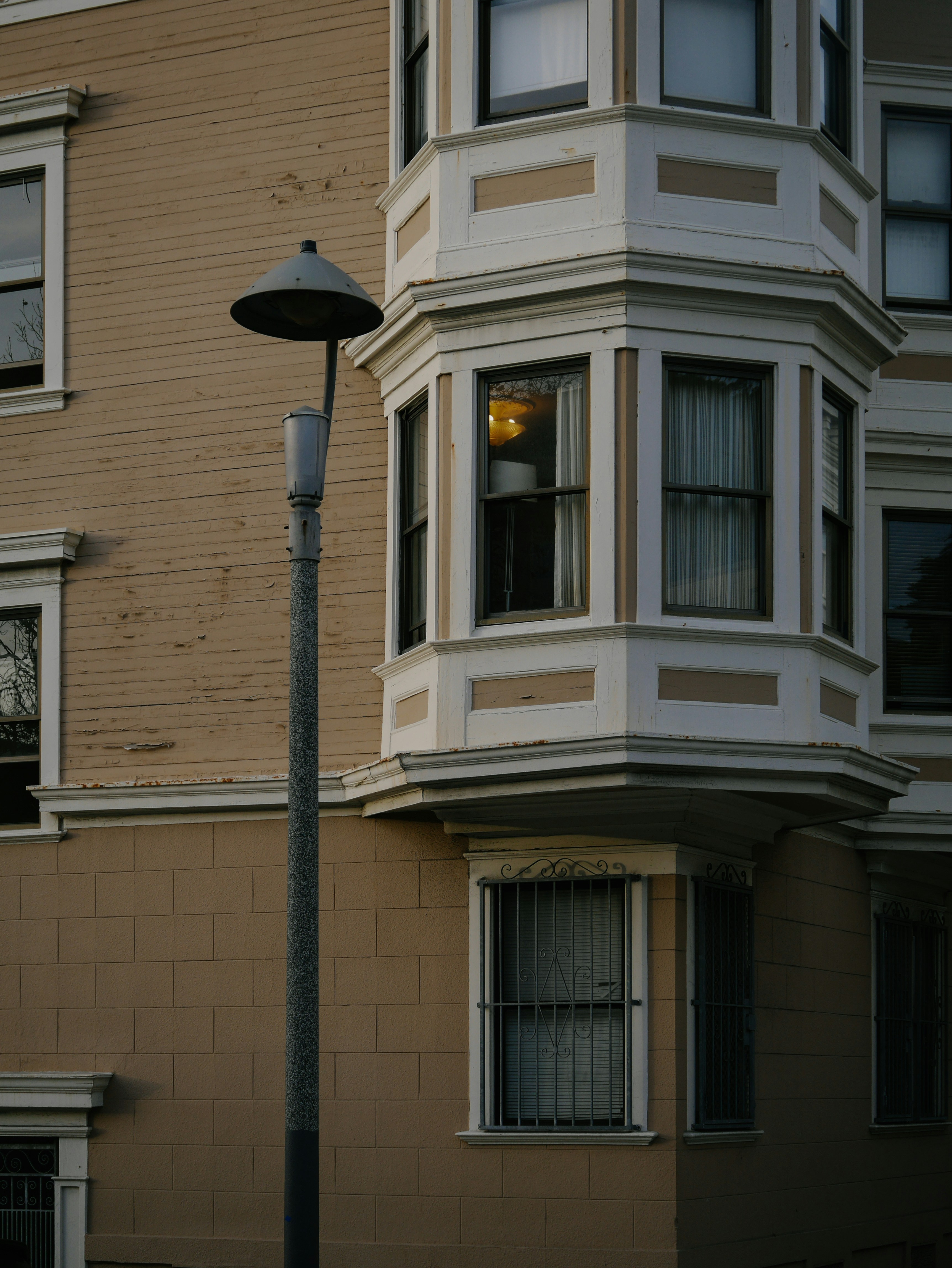 Warm light spills from a bay window on a pale beige building, with a lone streetlamp standing in the foreground. The composition emphasizes architectural detail and a quiet urban mood.