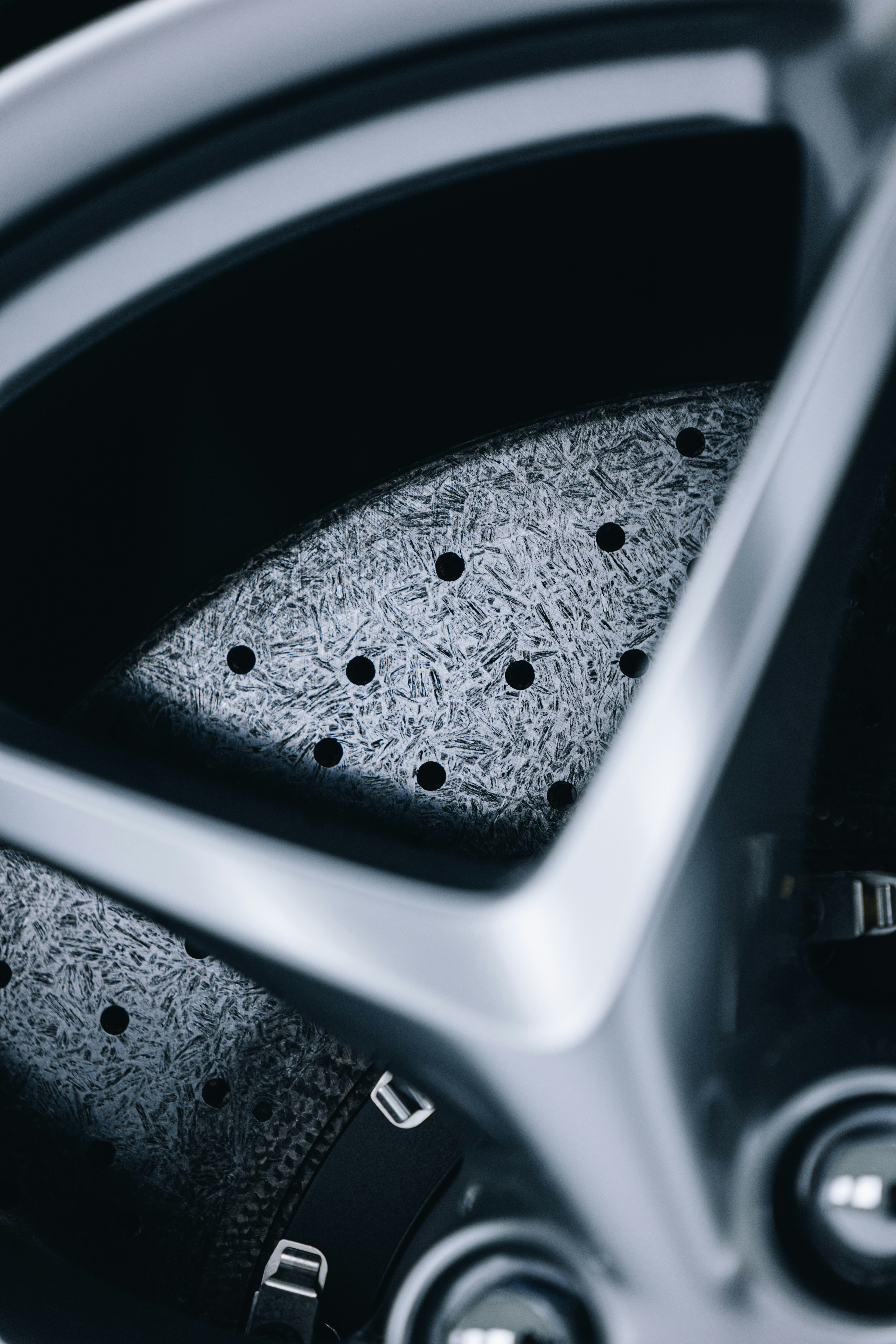 Close-up photograph of a perforated brake rotor inside a car wheel, illuminated in cool blue light. The textured metal surface and evenly spaced holes emphasize engineering precision.