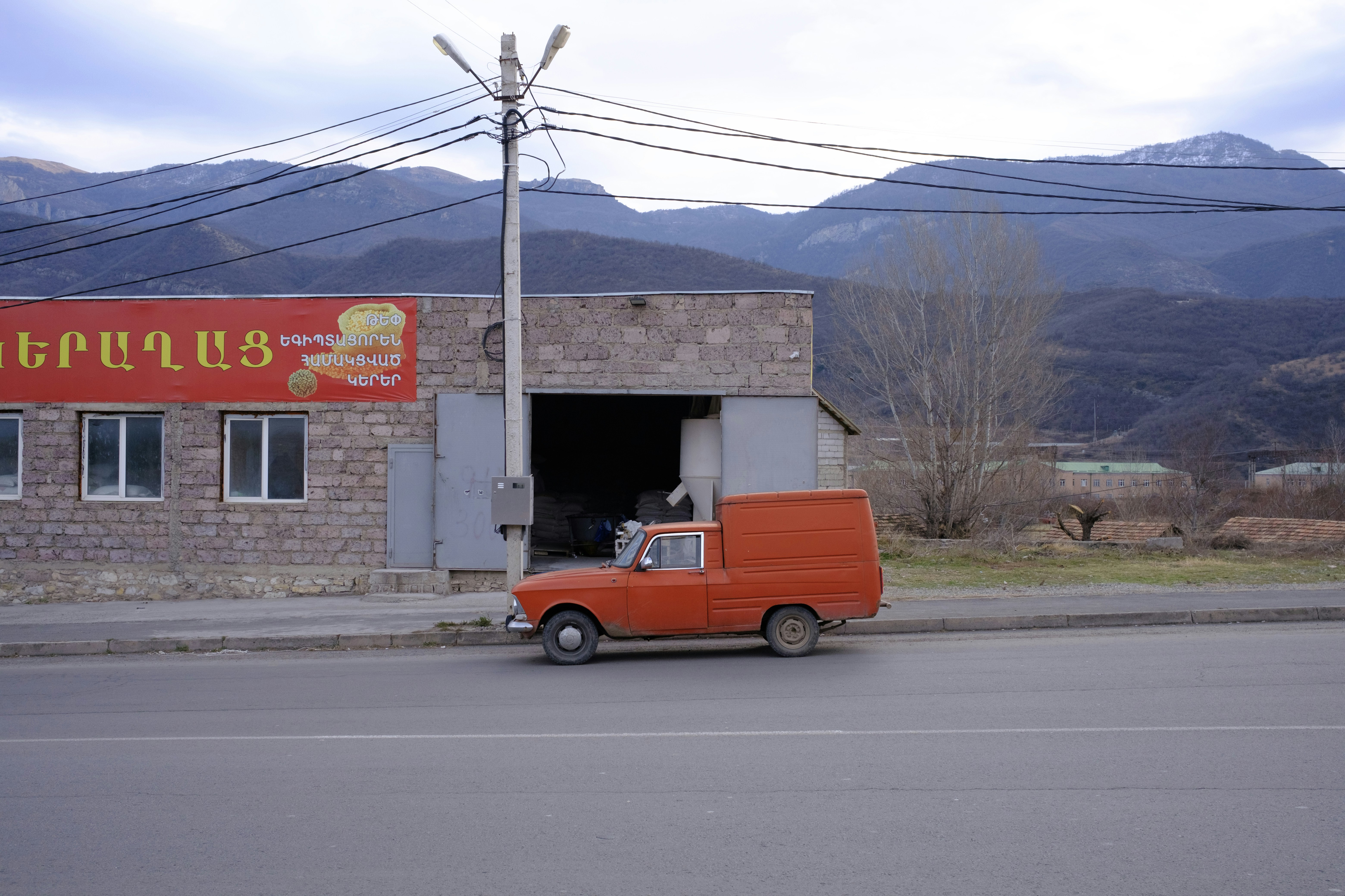 An orange van parked beside a stone building with a vibrant sign, set against a backdrop of mountains and a cloudy sky.