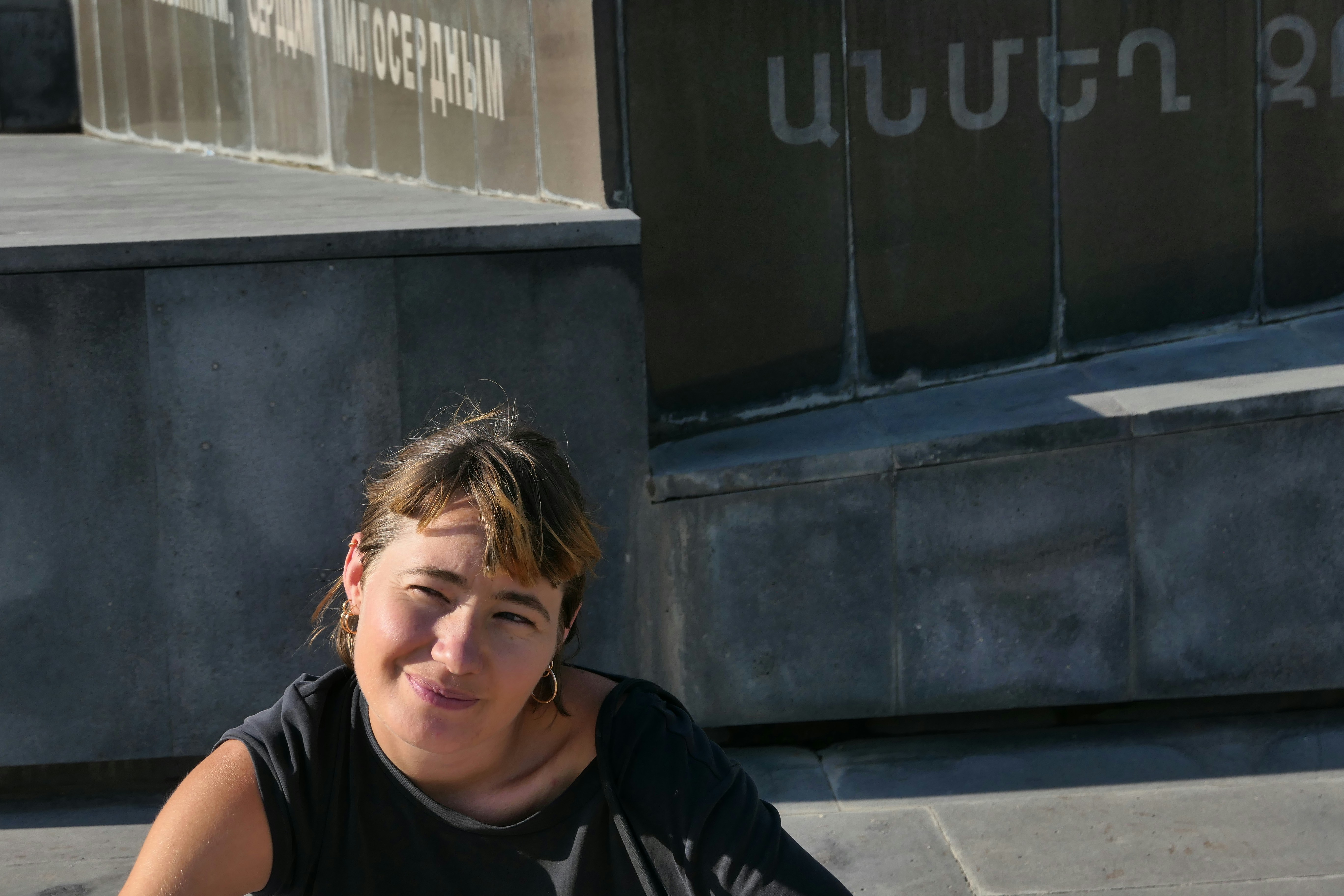 Woman seated in front of a concrete structure with engraved writing in sunlight.