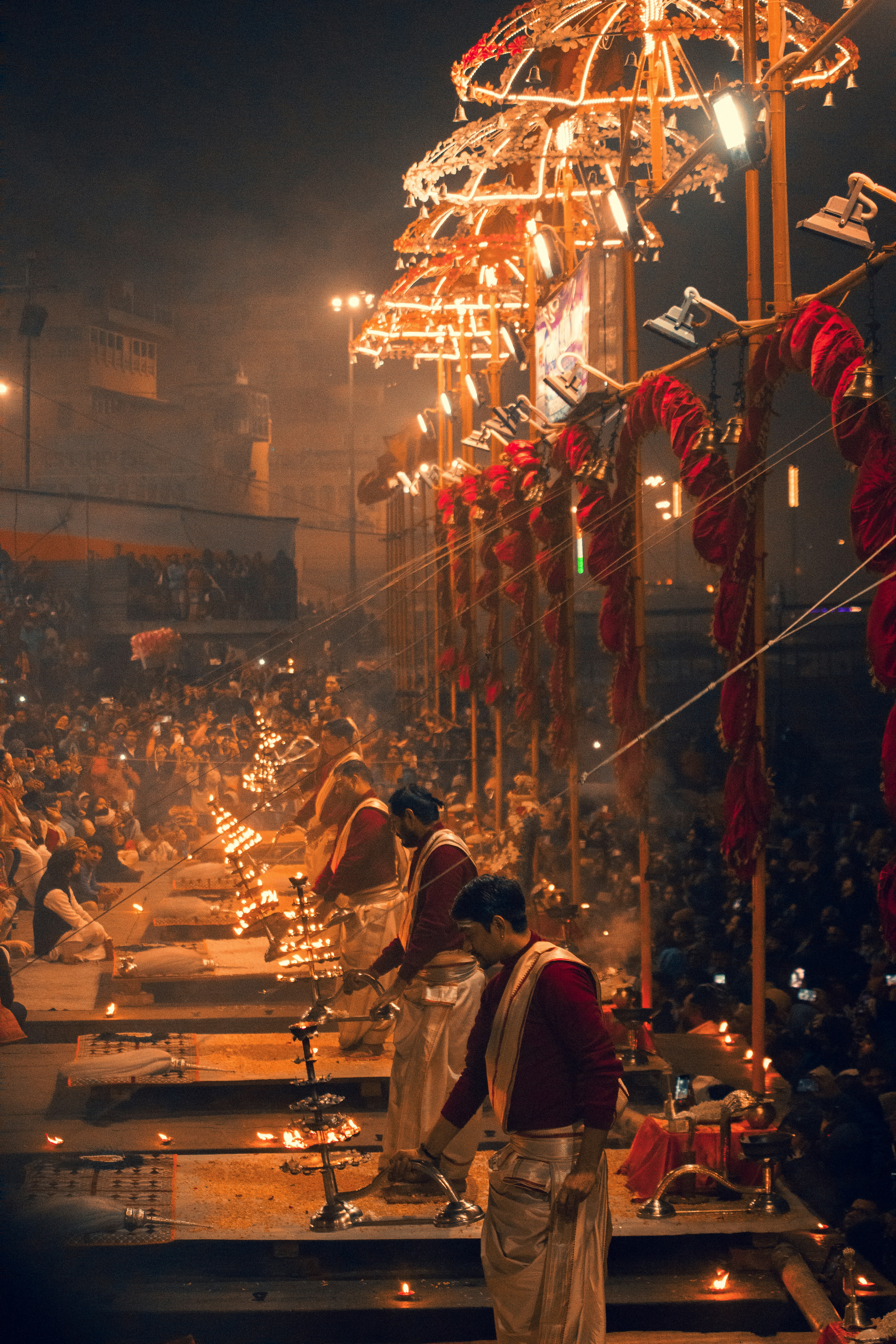 Hindu priests performing aarti with oil lamps along the banks of a river, surrounded by a captivated audience during a vibrant evening ceremony.