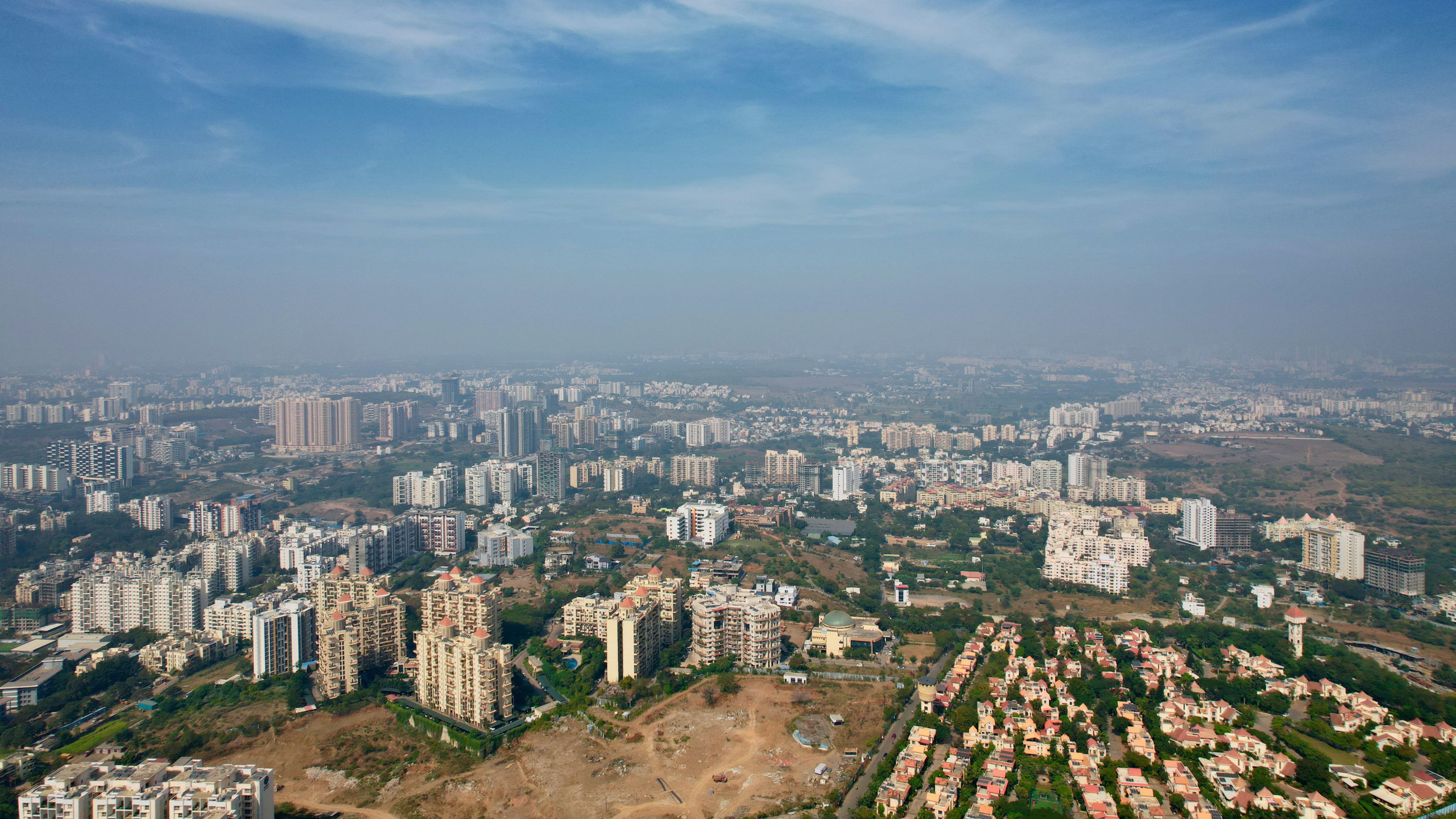This is a sky view of Undri, Pune, Maharashtra, India. Undri, a developing neighbourhood on the southern outskirts of Pune. Pune Metropolitan Region Development Authority has jurisdiction over it (PMRDA). Undri has a lot of vegetation cover and provides a tranquil setting for residential living. Additionally, it enjoys proximity to a number of essential employment hubs and sound connectivity. The majority of Undri comprises a variety of villas, plots, and multi-story apartment buildings. In this area, some projects are both finished and still under construction. | an aerial view of a city with lots of tall buildings