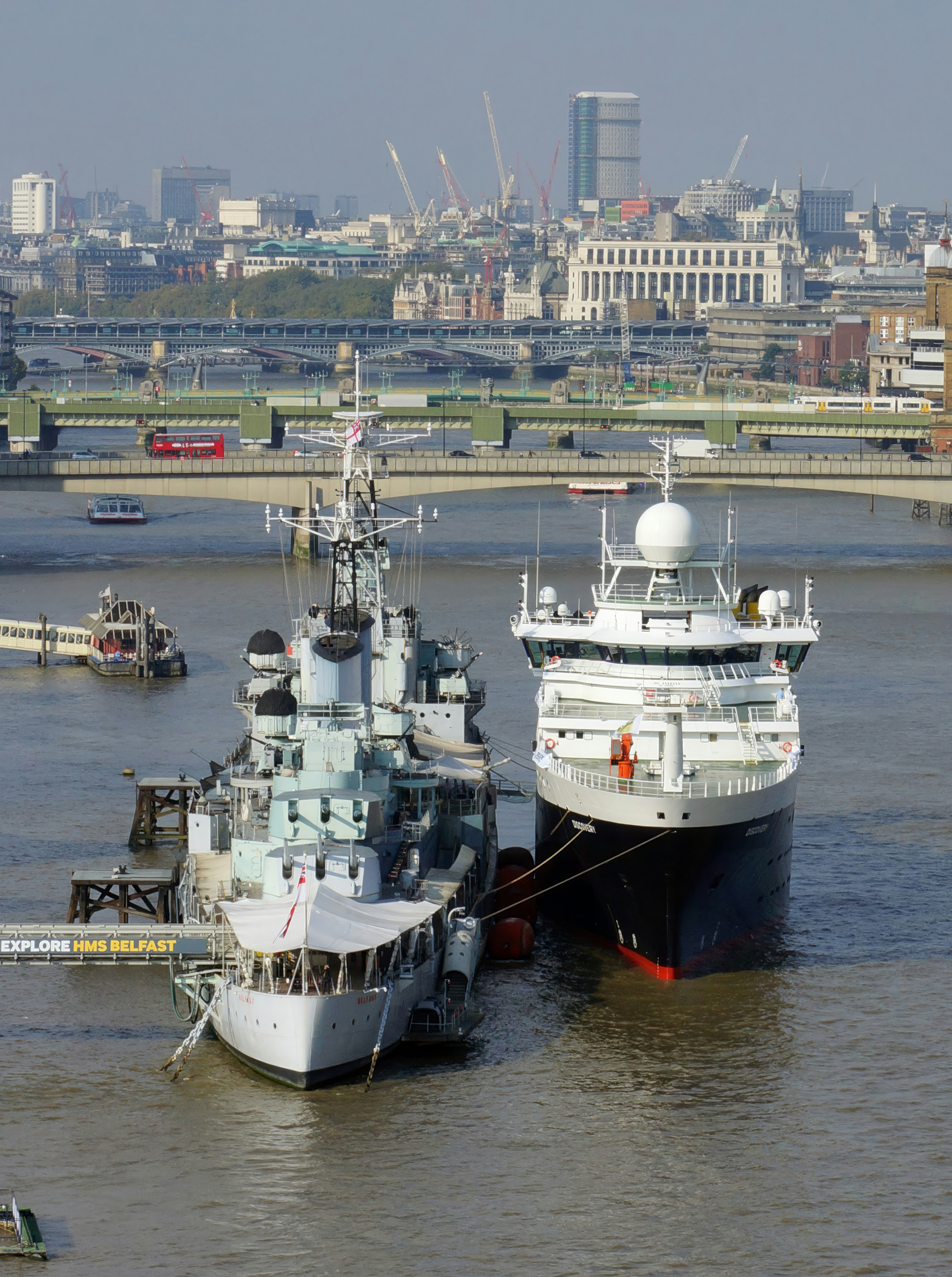 Two ships anchored side by side in the Thames, showcasing historical and modern maritime design against a bustling city backdrop.