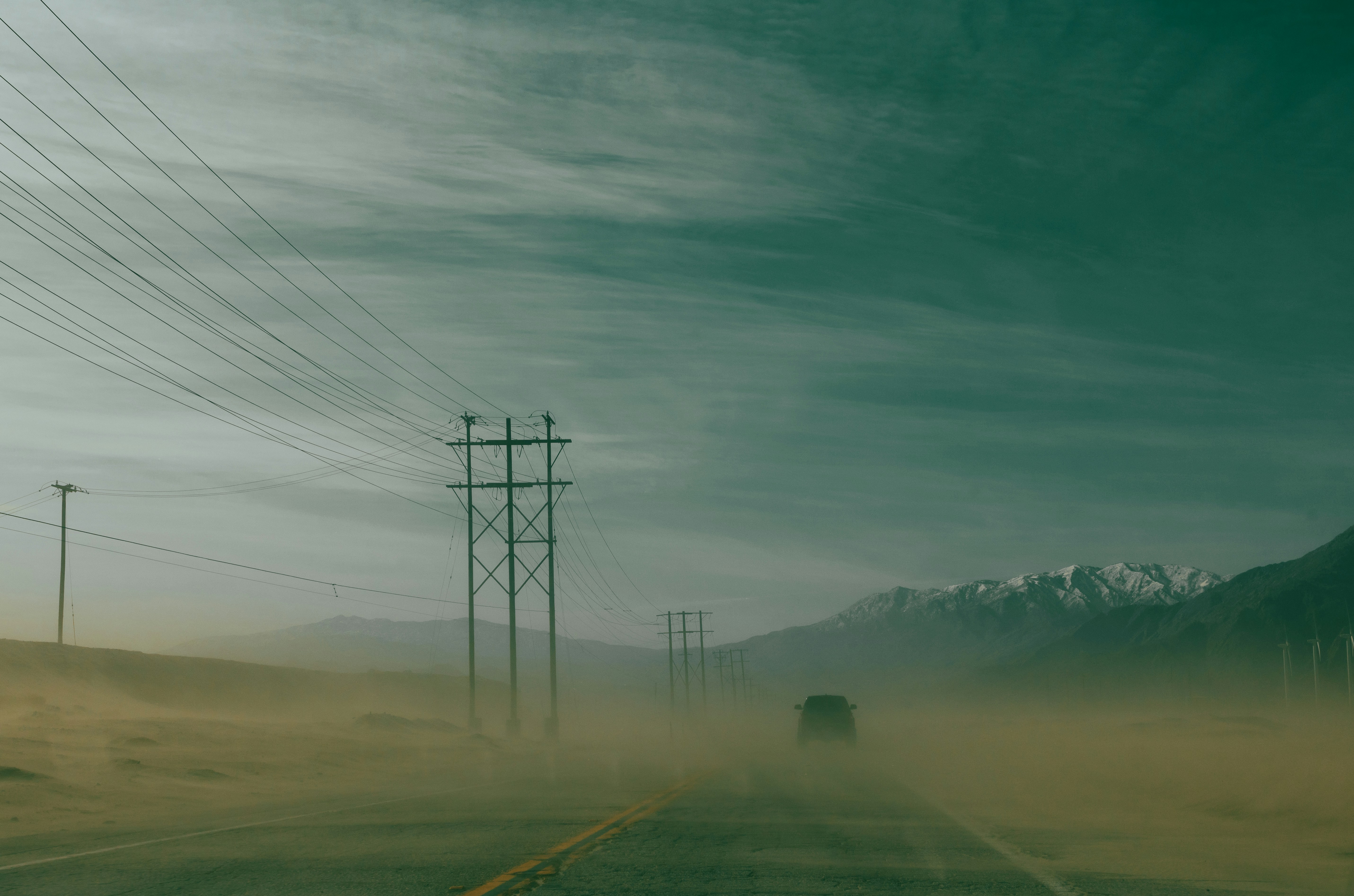 Car traveling along a desolate desert road with distant mountains and power lines.