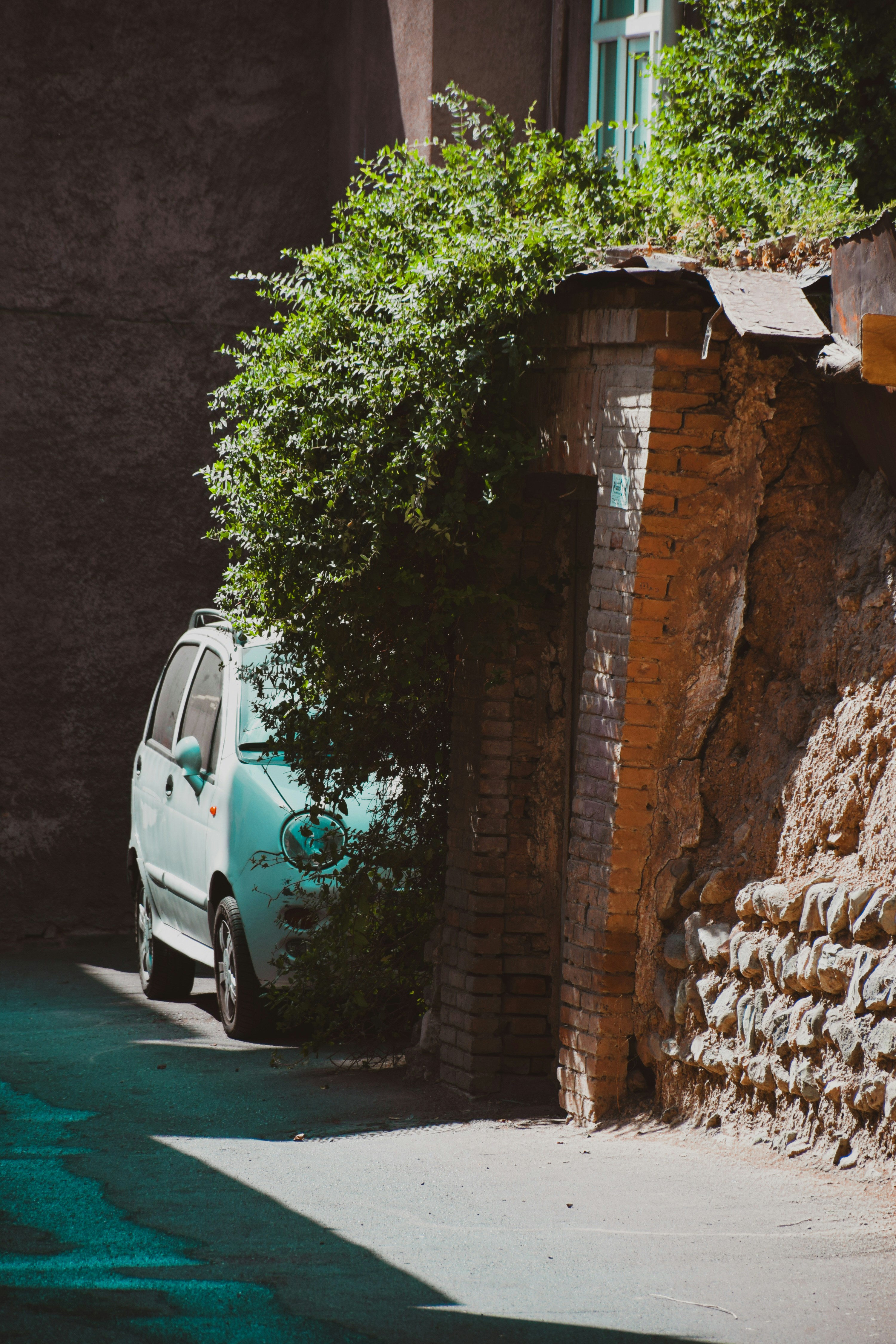 Hidden teal car tucked beside sunlit brick wall