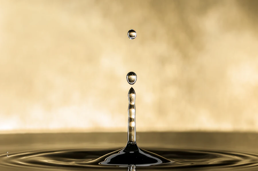 A single water droplet hangs above a glossy splash peak, with warm golden reflections and soft ripples spreading across the surface.