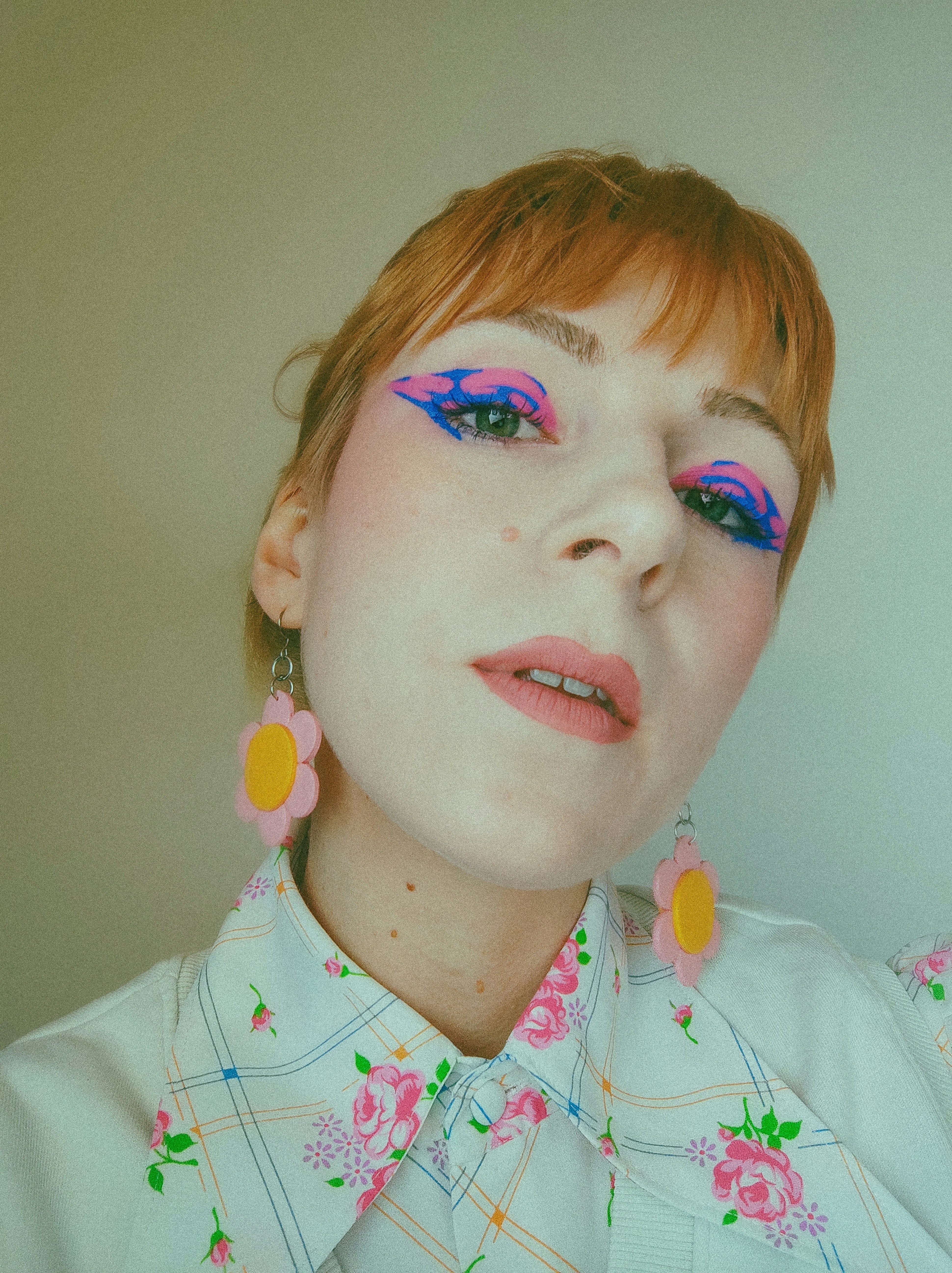 Woman with vivid eye makeup and floral earrings gazes confidently, wearing a floral-patterned shirt.