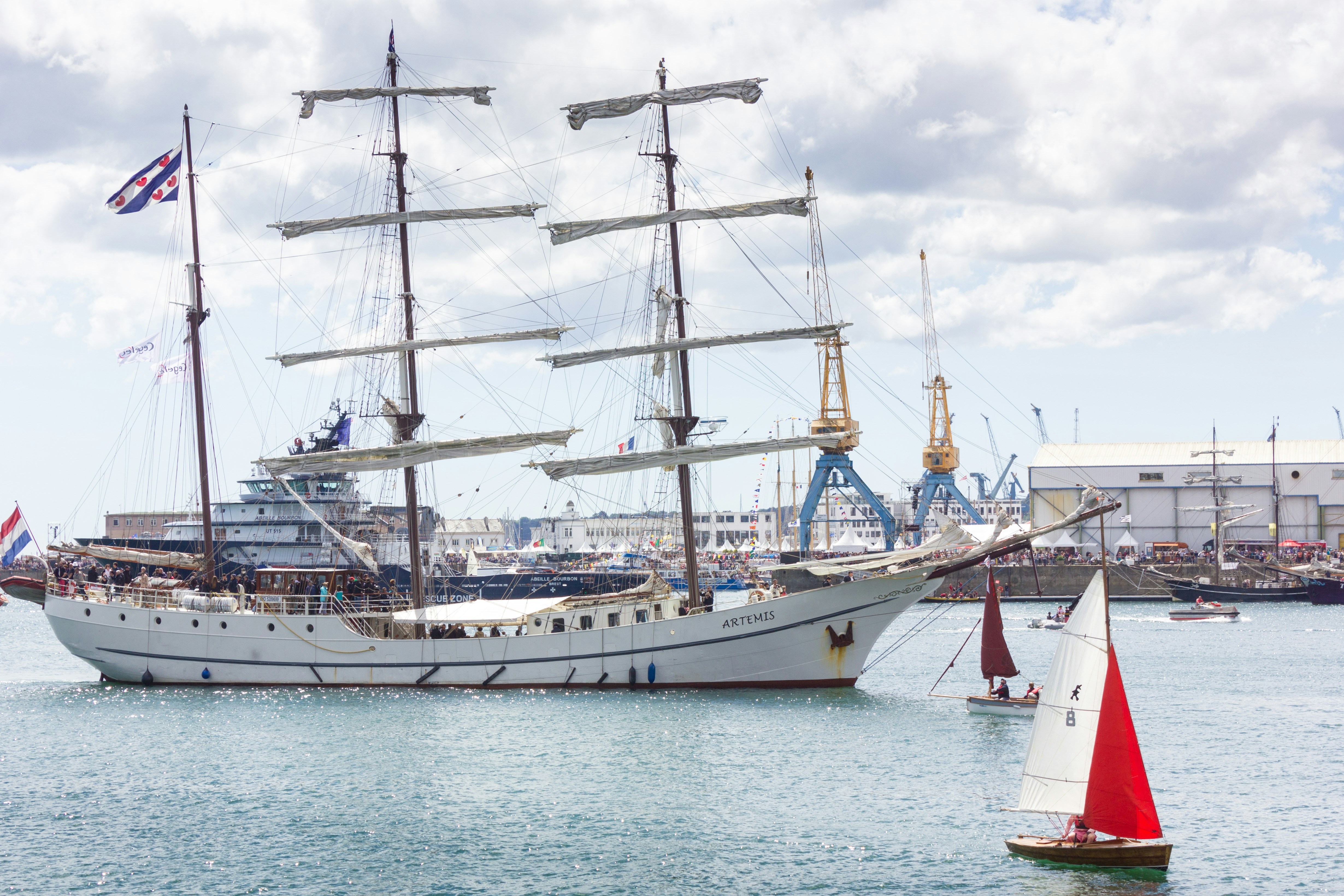 Tall ship Artemis sails gracefully in the harbor alongside smaller vessels under a partly cloudy sky.