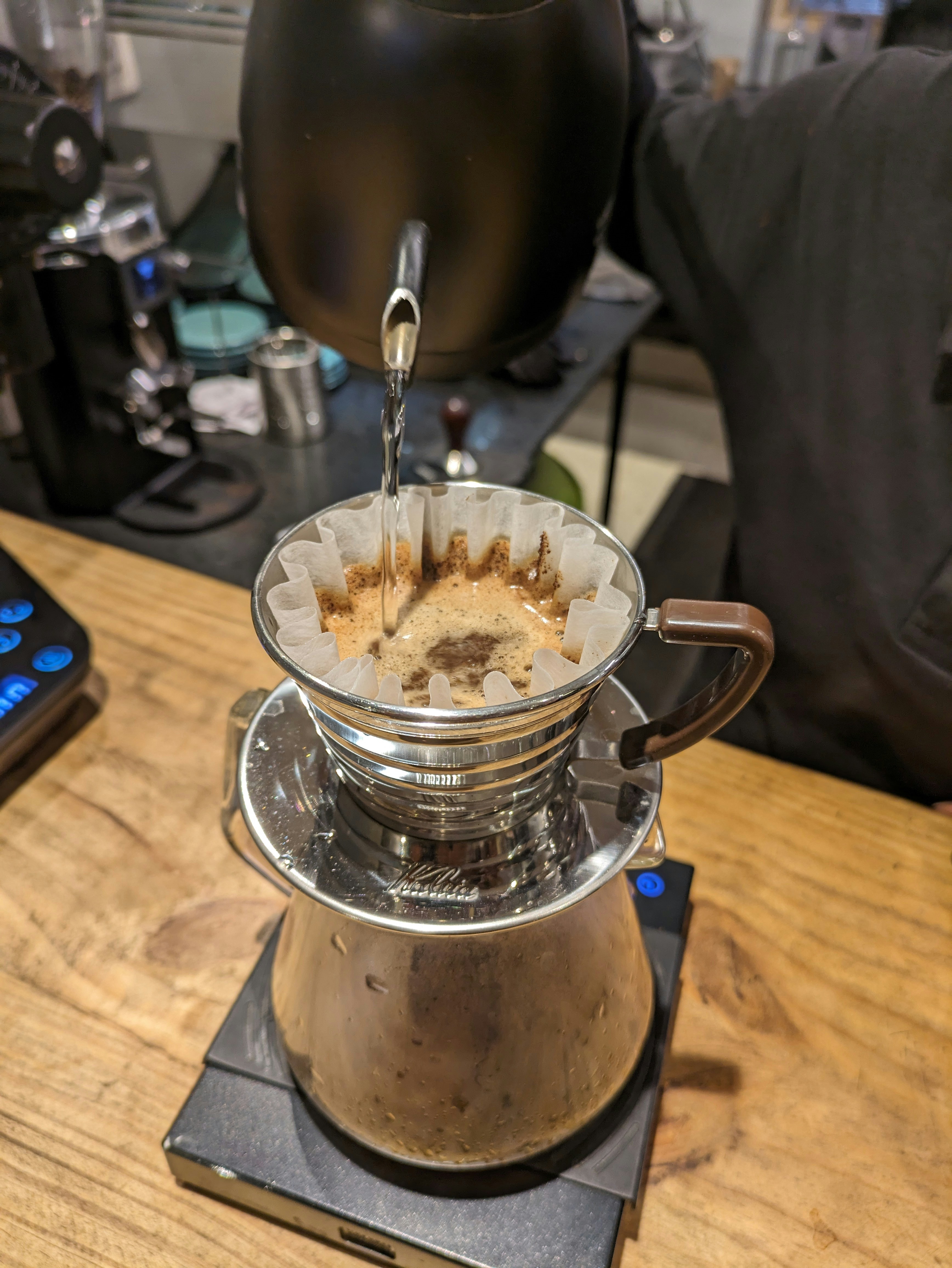 Pour-over setup with a paper filter in a metal dripper sits atop a precise scale while water blooms through the coffee grounds. The chrome kettle pours above a wood-toned counter with blurred café equipment in the background.