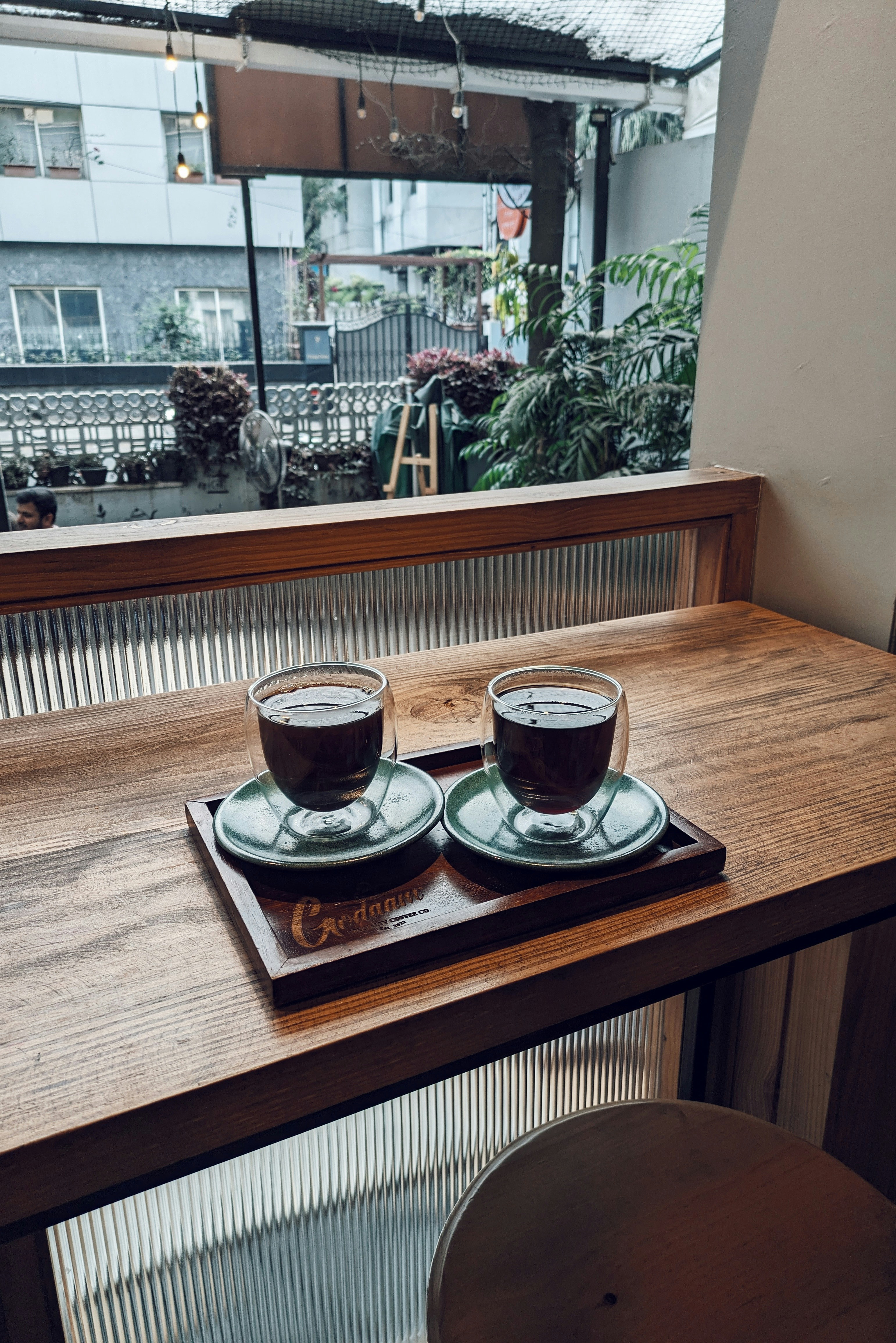 Two cups of dark coffee sit on a wooden tray along a cafe counter, framed by a sunlit window and lush greenery outside.