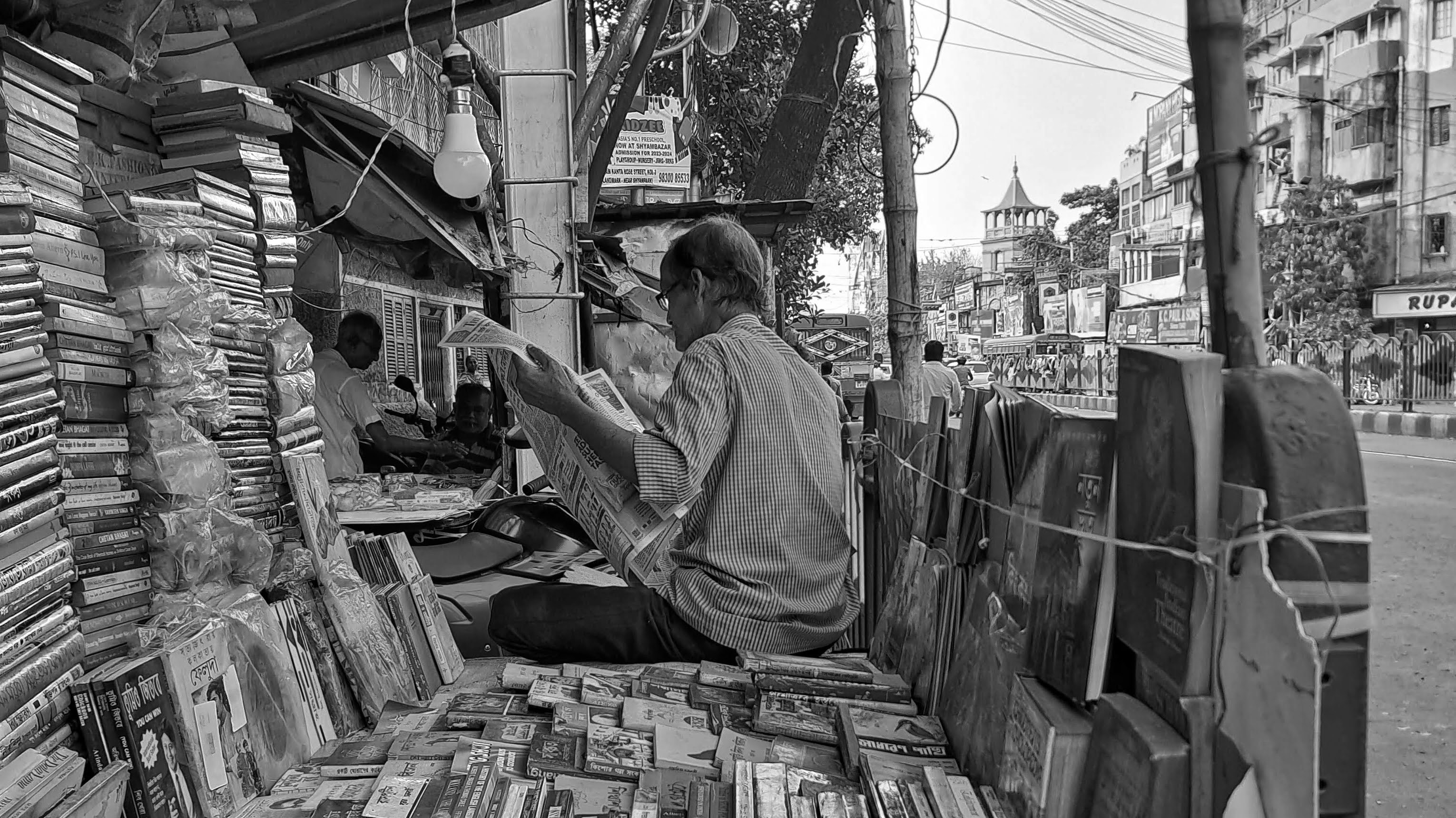 Black and white image of a bookseller surrounded by stacks of books in an outdoor stall on a busy street.
