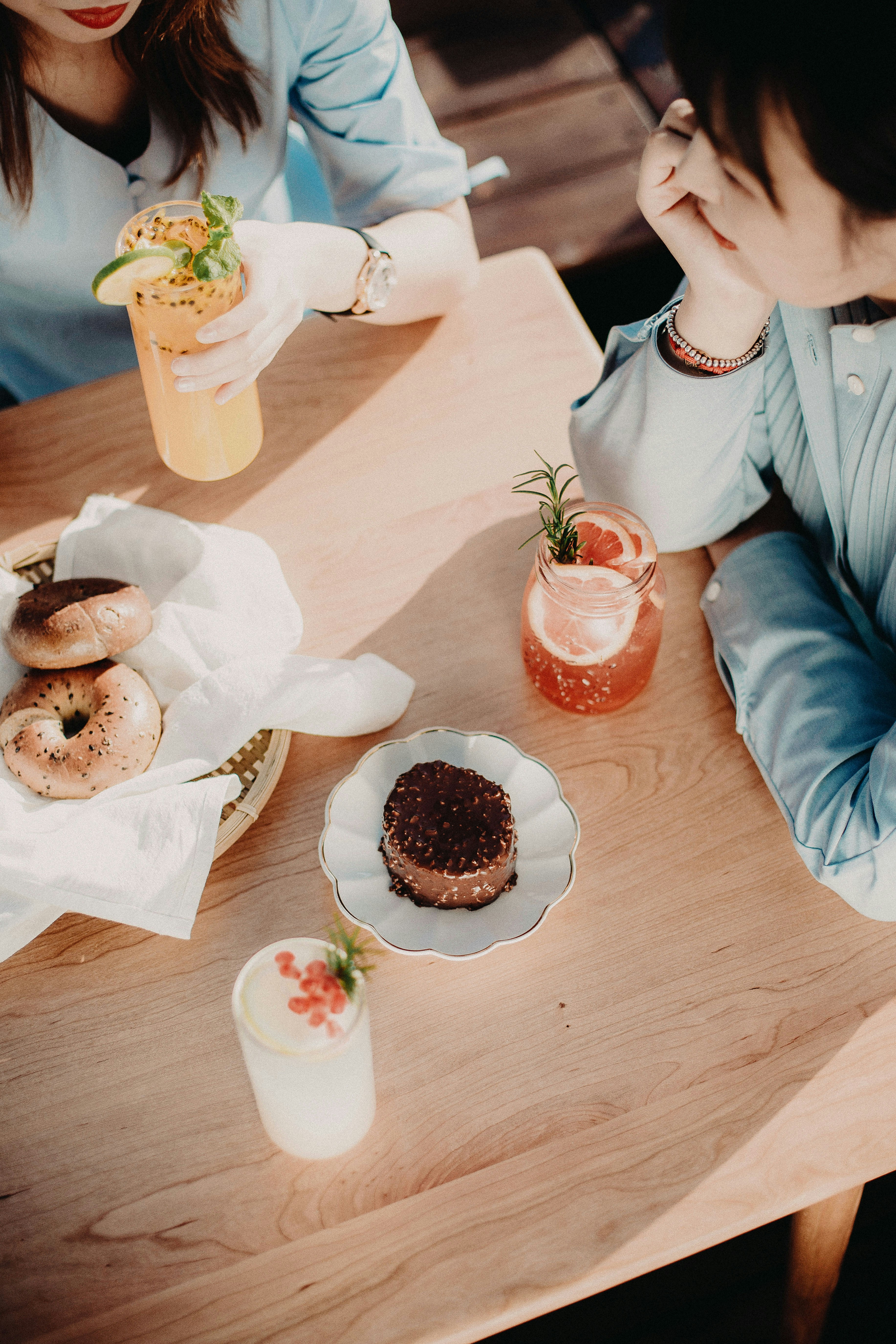 A cozy table setting featuring an assortment of beverages and treats, including bagels, a chocolate dessert, and colorful cocktails. Friends enjoy their time together over delightful snacks.