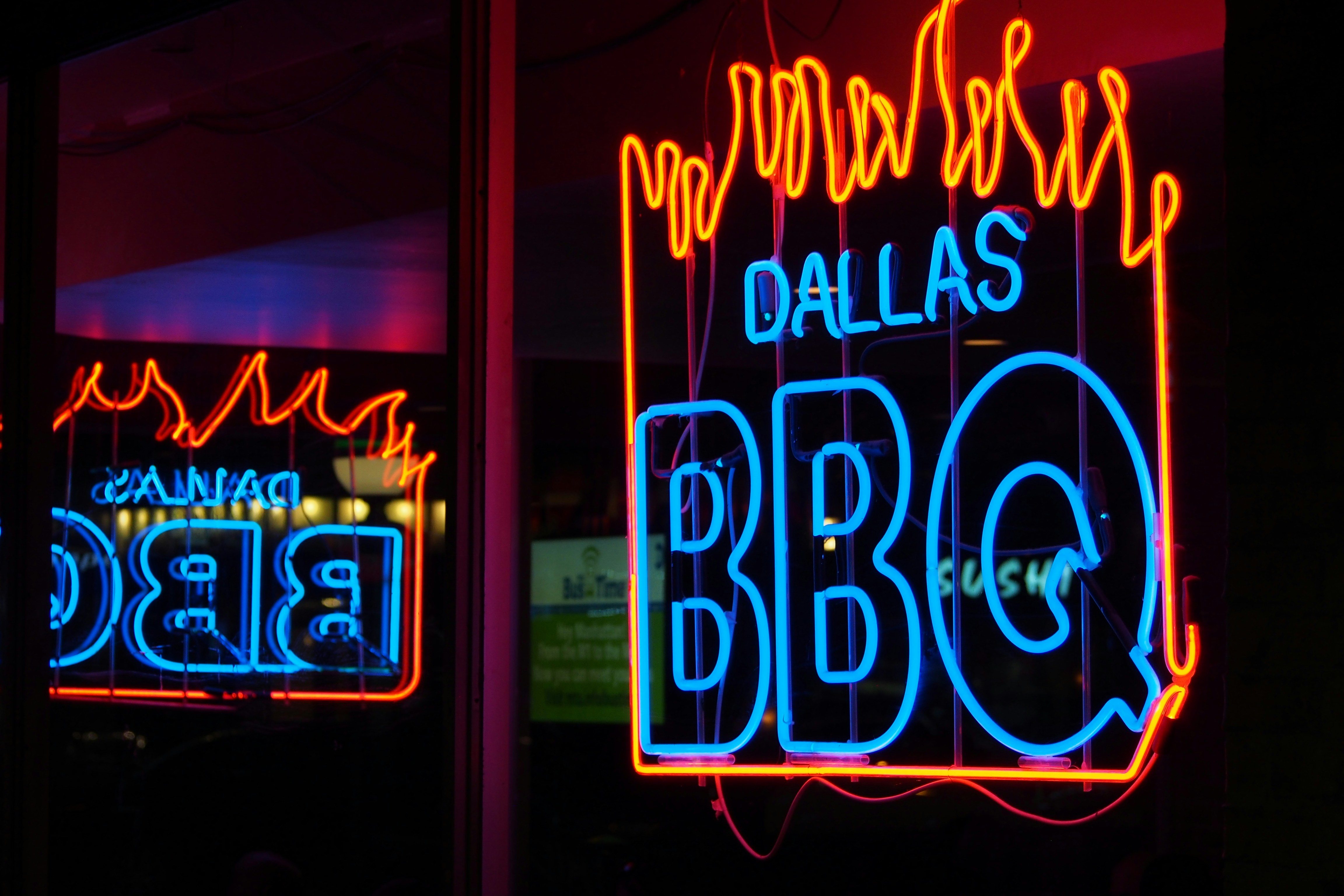 Blue neon 'BBQ' with orange accents spell 'DALLAS' in a storefront window, with the glow mirrored on the glass.