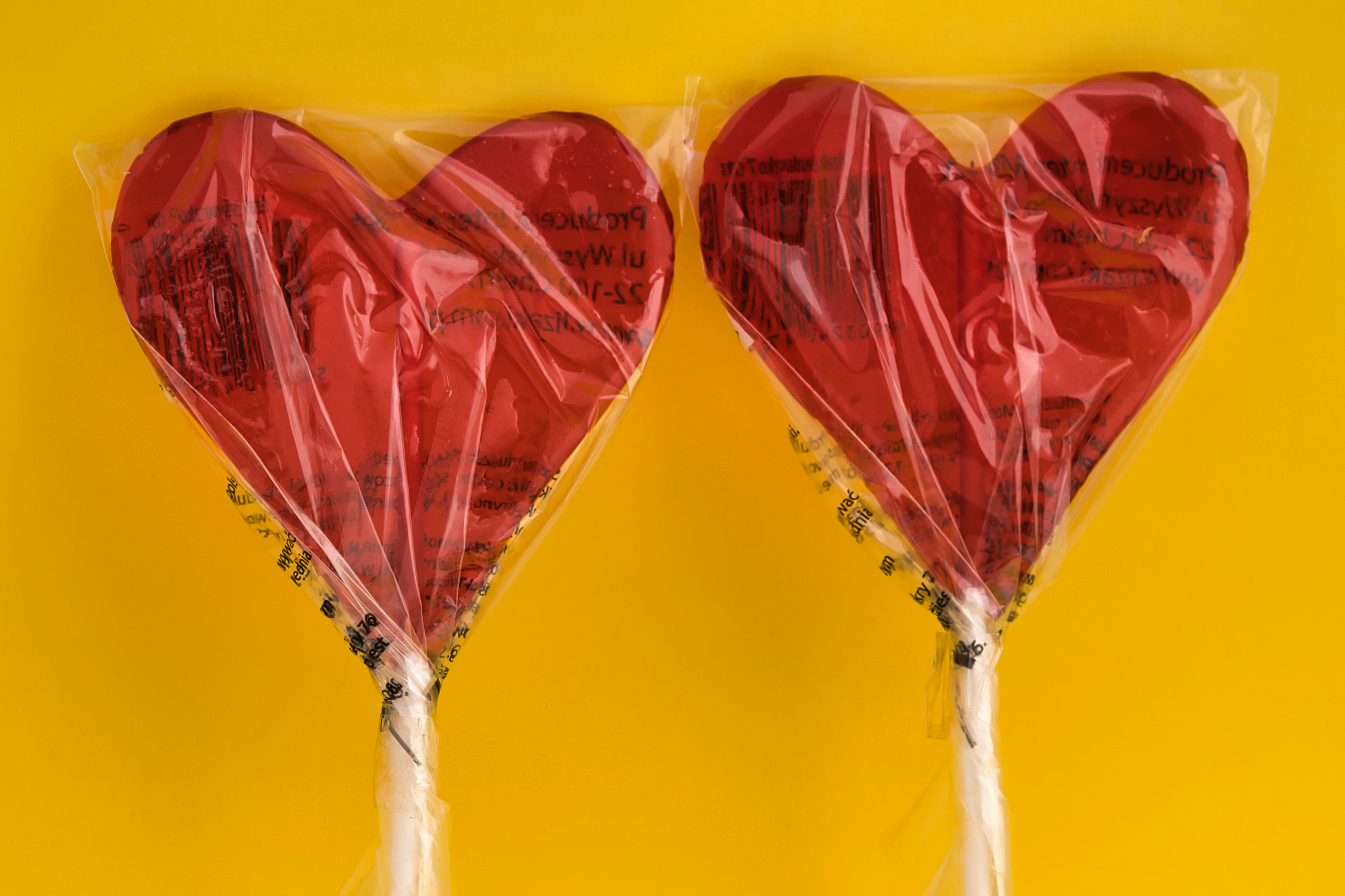 Two heart-shaped lollipops wrapped in clear plastic against a vibrant yellow background.