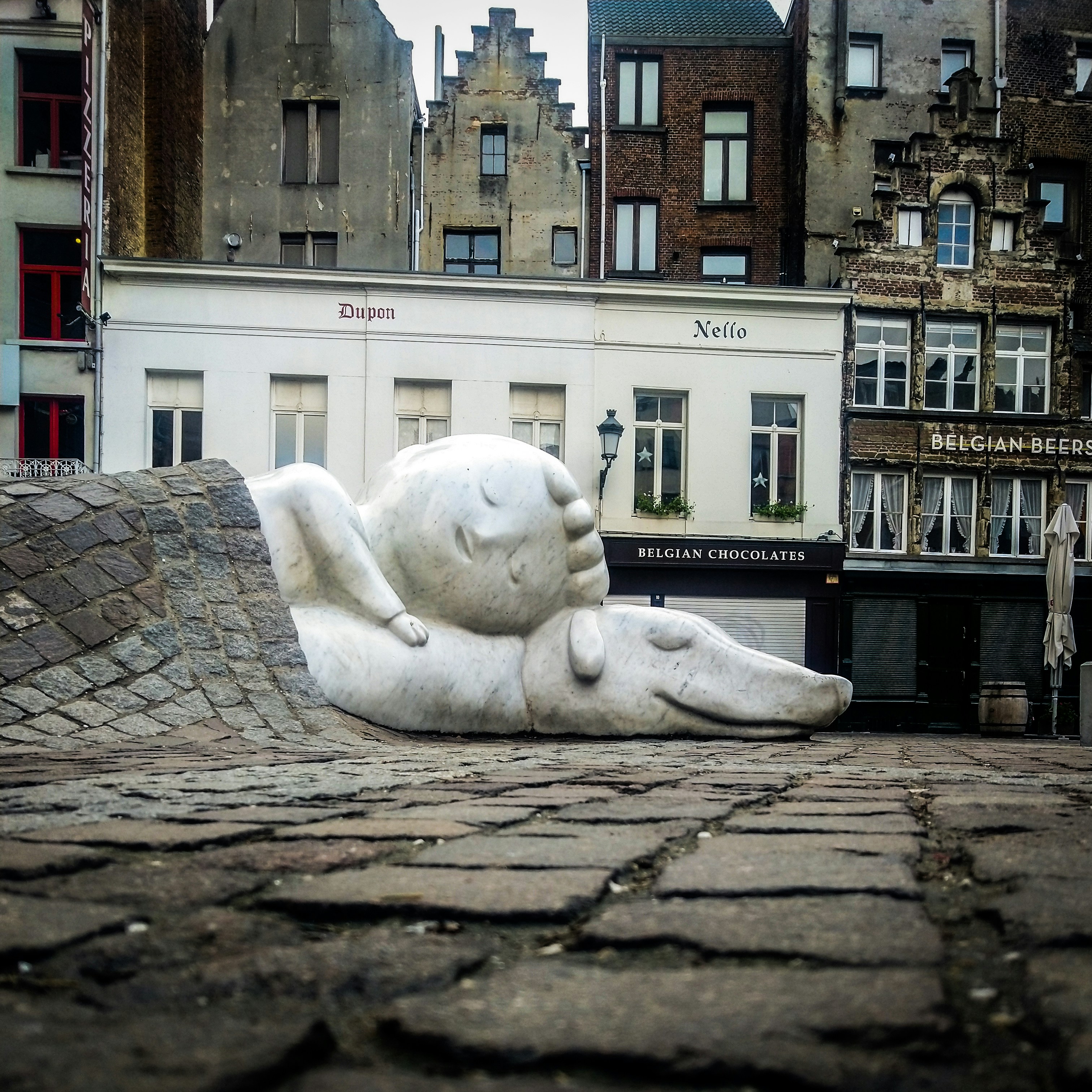 Colossal white reclining sculpture rests on a cobbled square, with historic brick storefronts and a Belgian chocolates shop in the background.