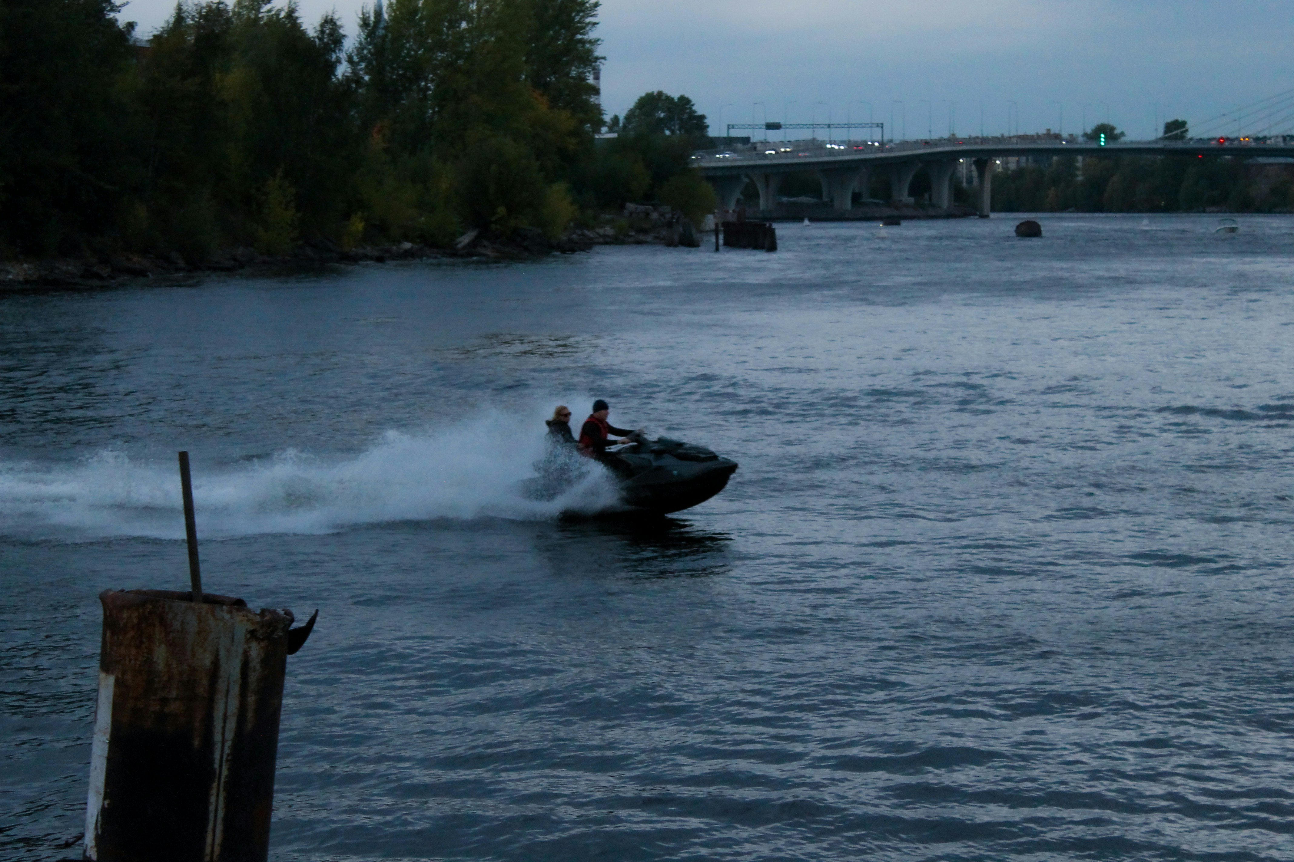 Jet ski racing across a river at dusk, with a bridge visible in the background. The scene captures the excitement of water sports under the fading light.