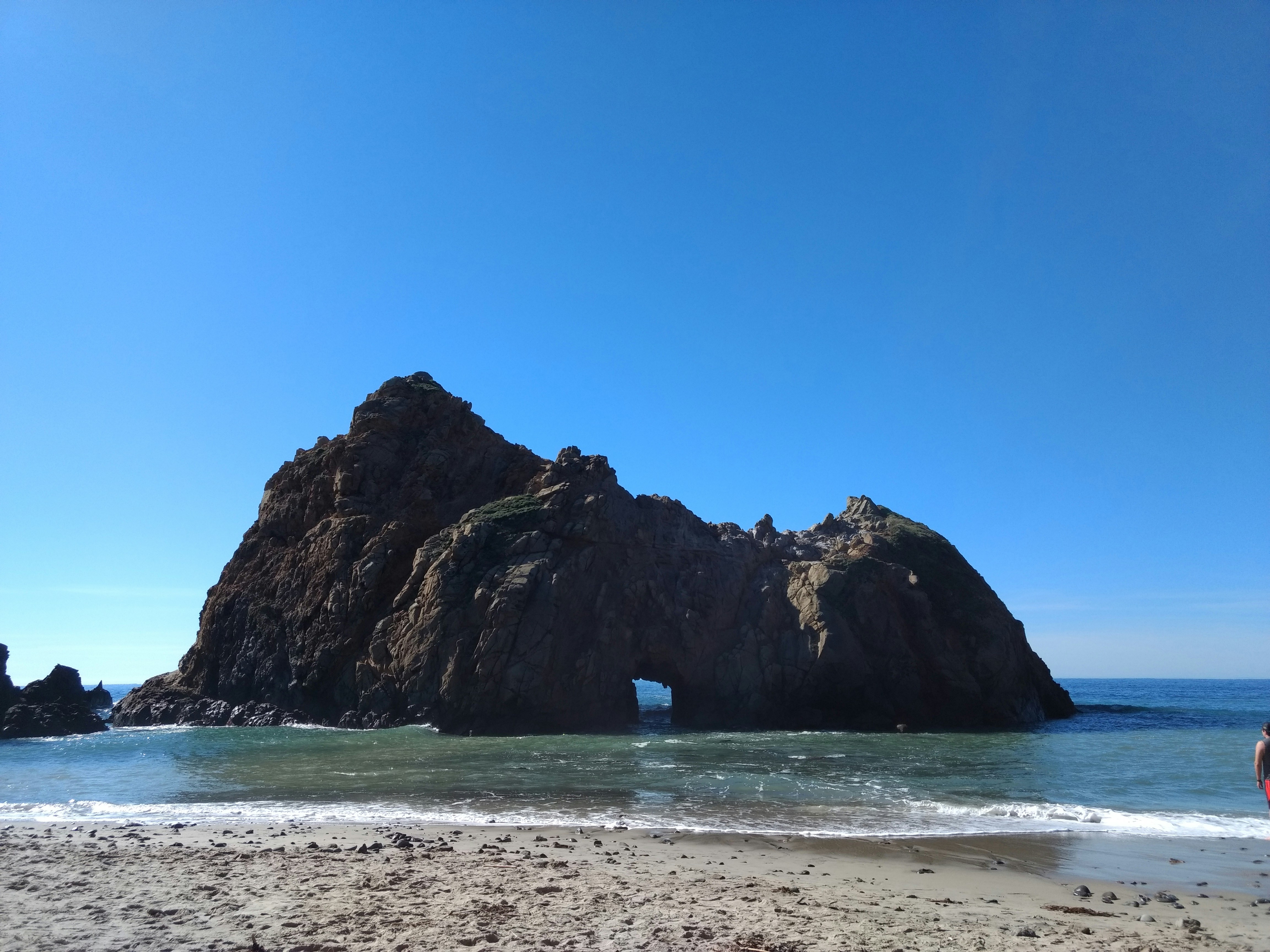 Photograph of a rugged sea arch rising from a calm shoreline, framed by a vivid blue sky and gentle surf.
